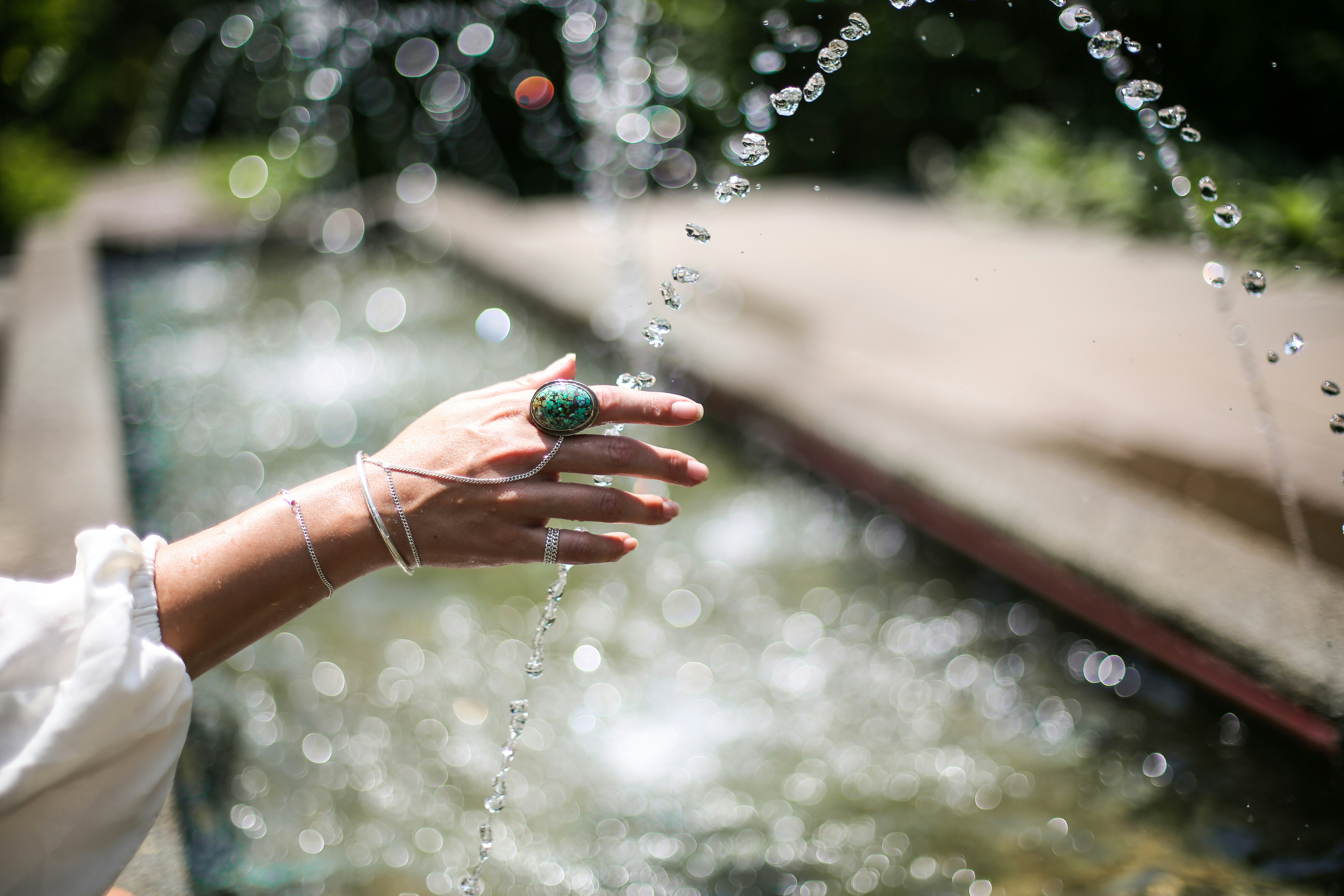Hand adorned with rings gracefully poised above a water fountain, capturing the motion of droplets in sunlight.