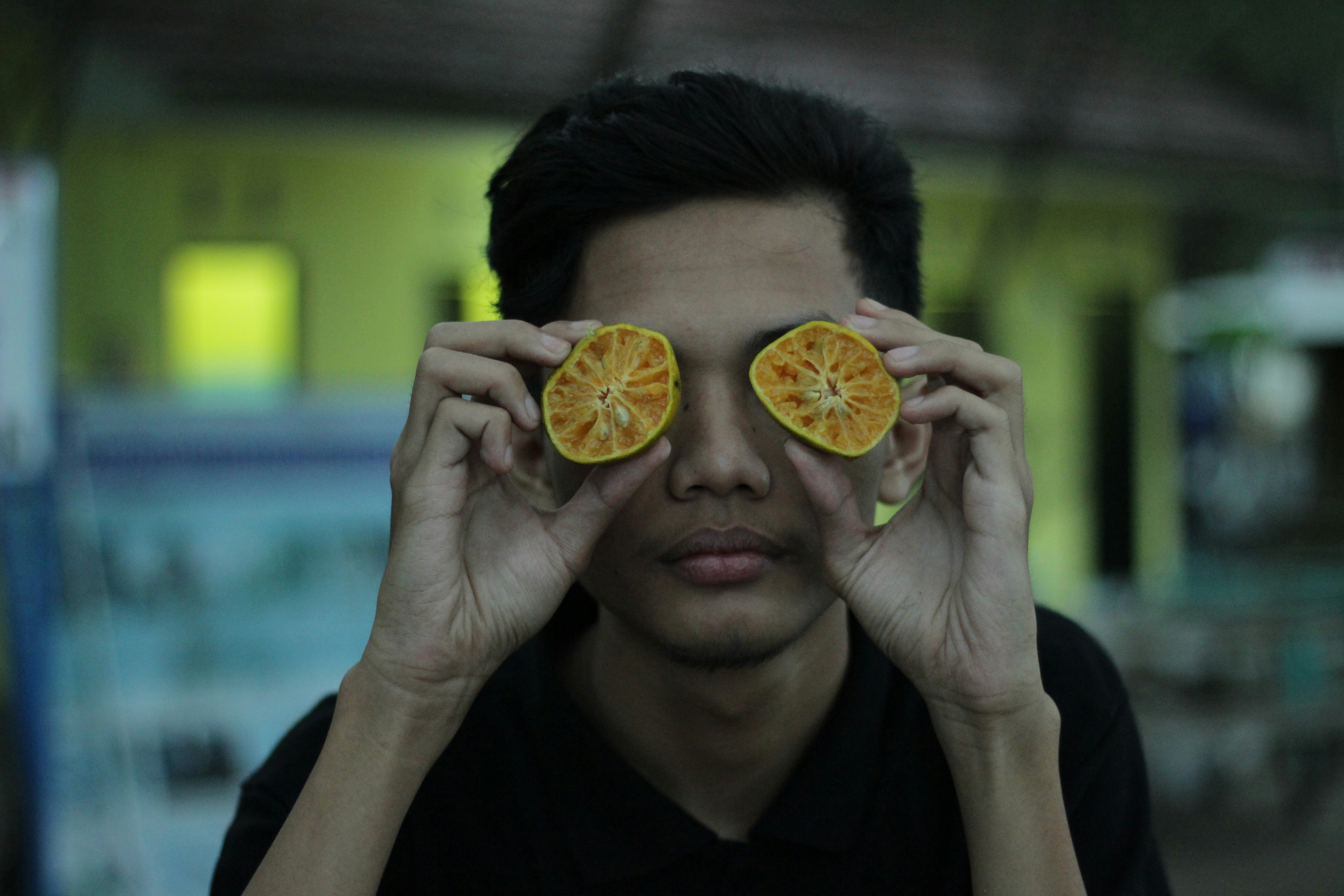 Young man holding orange halves in front of his eyes, creating a whimsical expression. The background features soft, blurred lights suggesting an evening setting.
