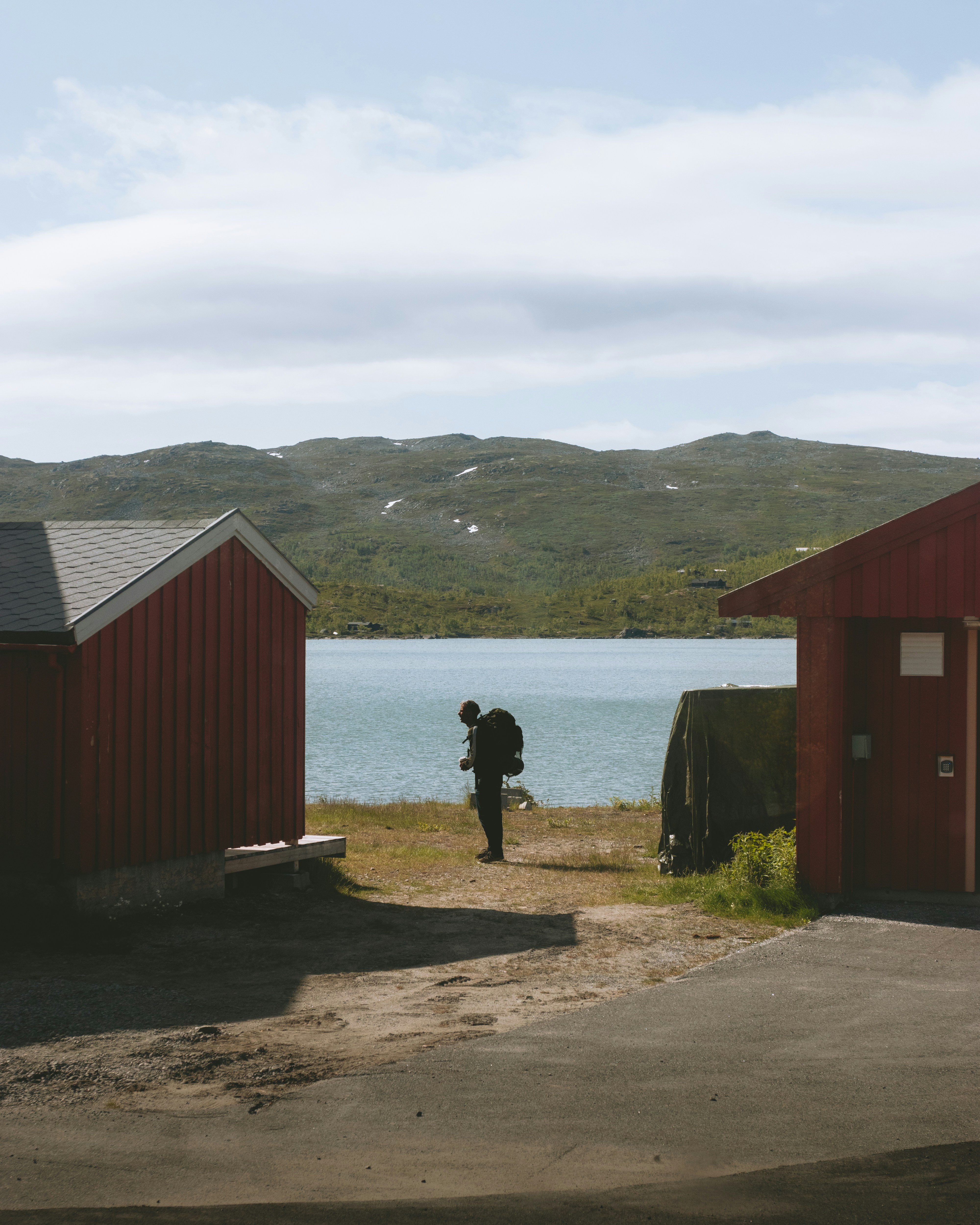 A lone hiker with a backpack stands near a serene lake, flanked by rustic red cabins and lush green hills in the background.