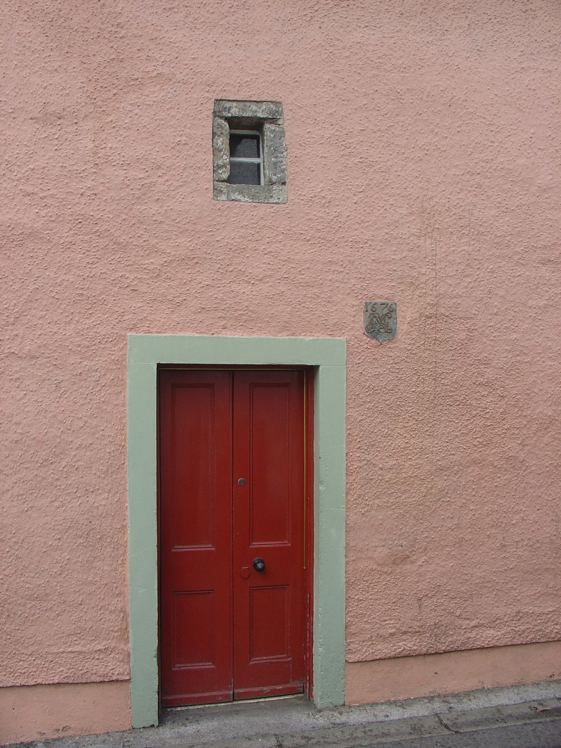 A vibrant red double door framed by a light green border, set against a textured pink wall, with a small stone window above and a year plaque to the side.