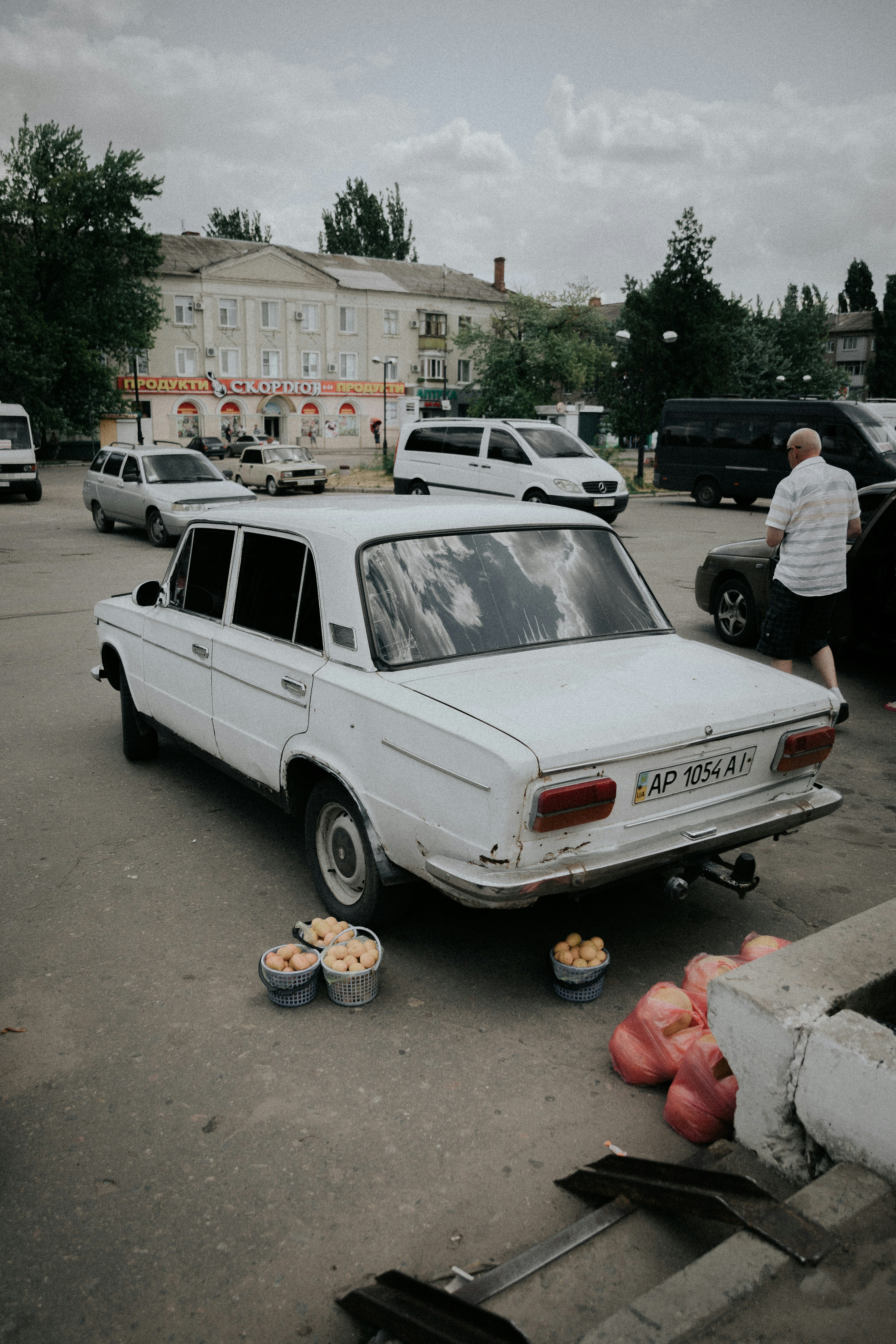 A vintage white car parked in a bustling market area, surrounded by baskets of fresh produce and a few pedestrians. The scene captures everyday life in a city.