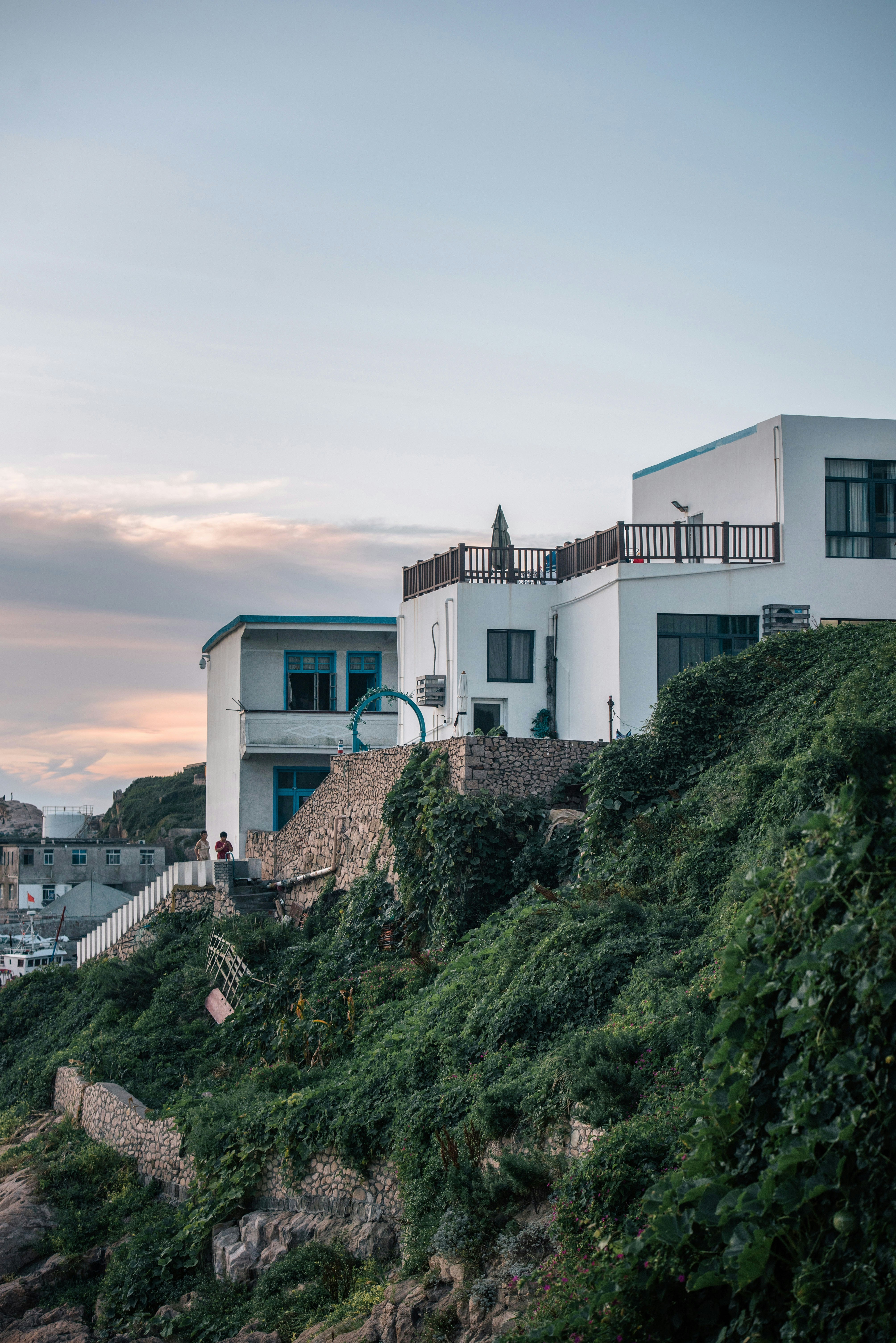 A contemporary building nestled on a lush hillside, overlooking the sea, with a sunset sky in the background.