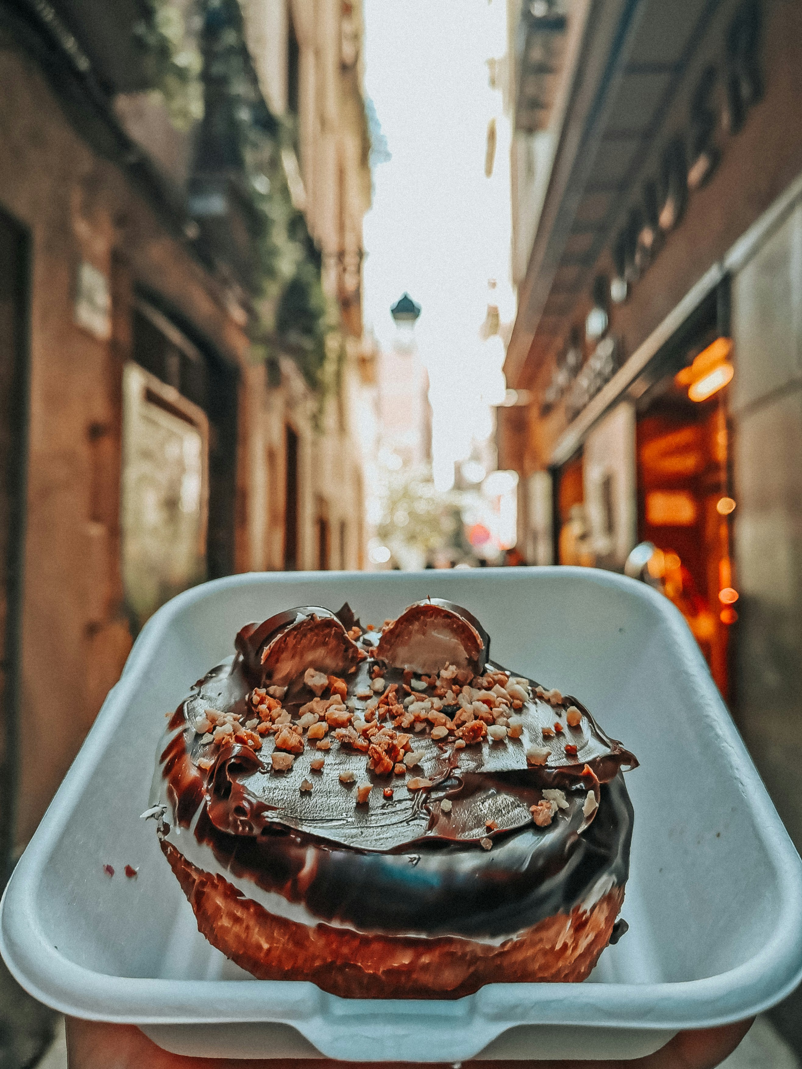 Decadent chocolate-covered donut topped with crushed nuts, held in a white container against a sunlit alley backdrop.