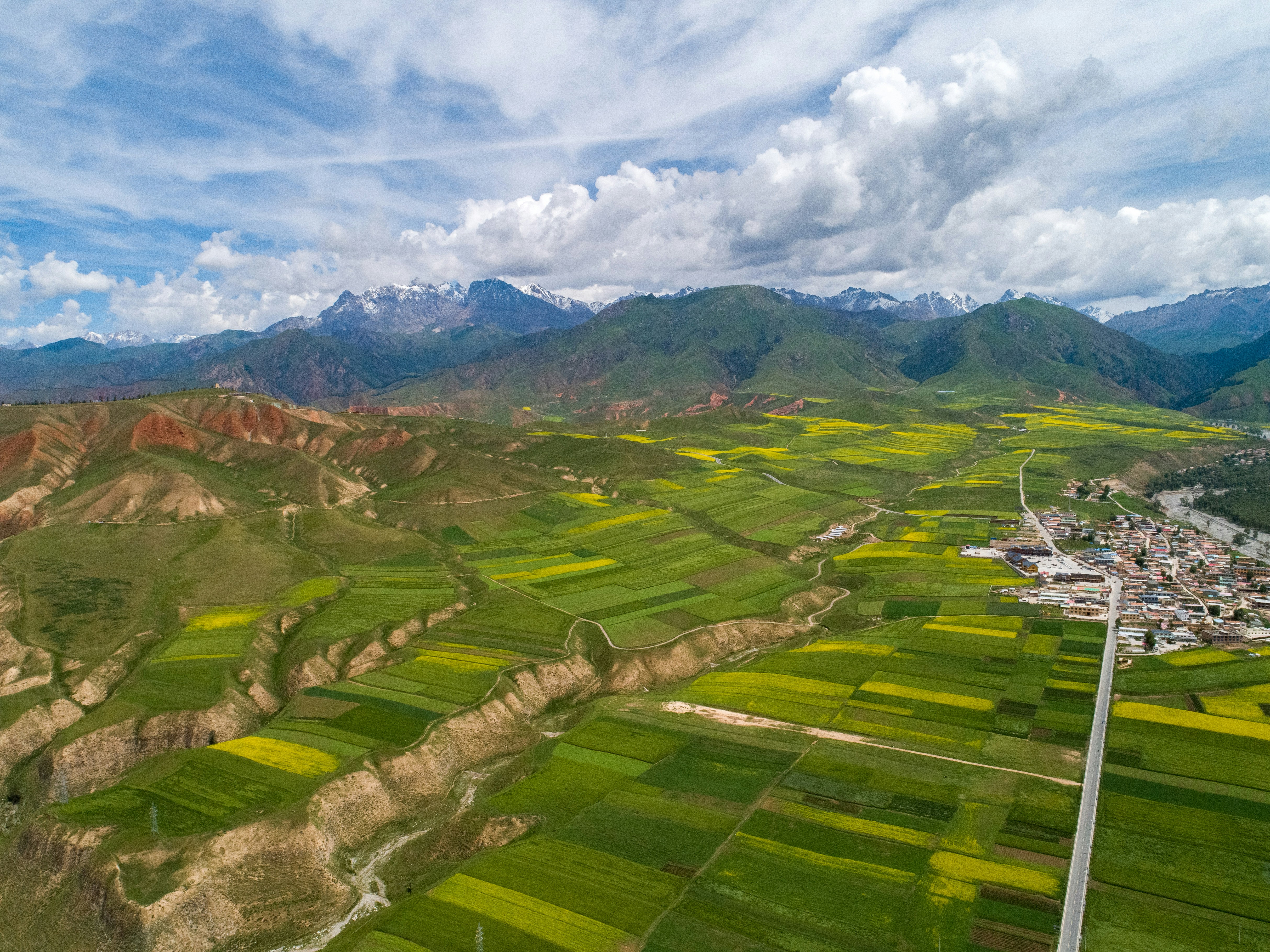 Expansive fields in varying shades of green and yellow stretch across the landscape, bordered by rugged mountains and a small town nestled at the edge. 