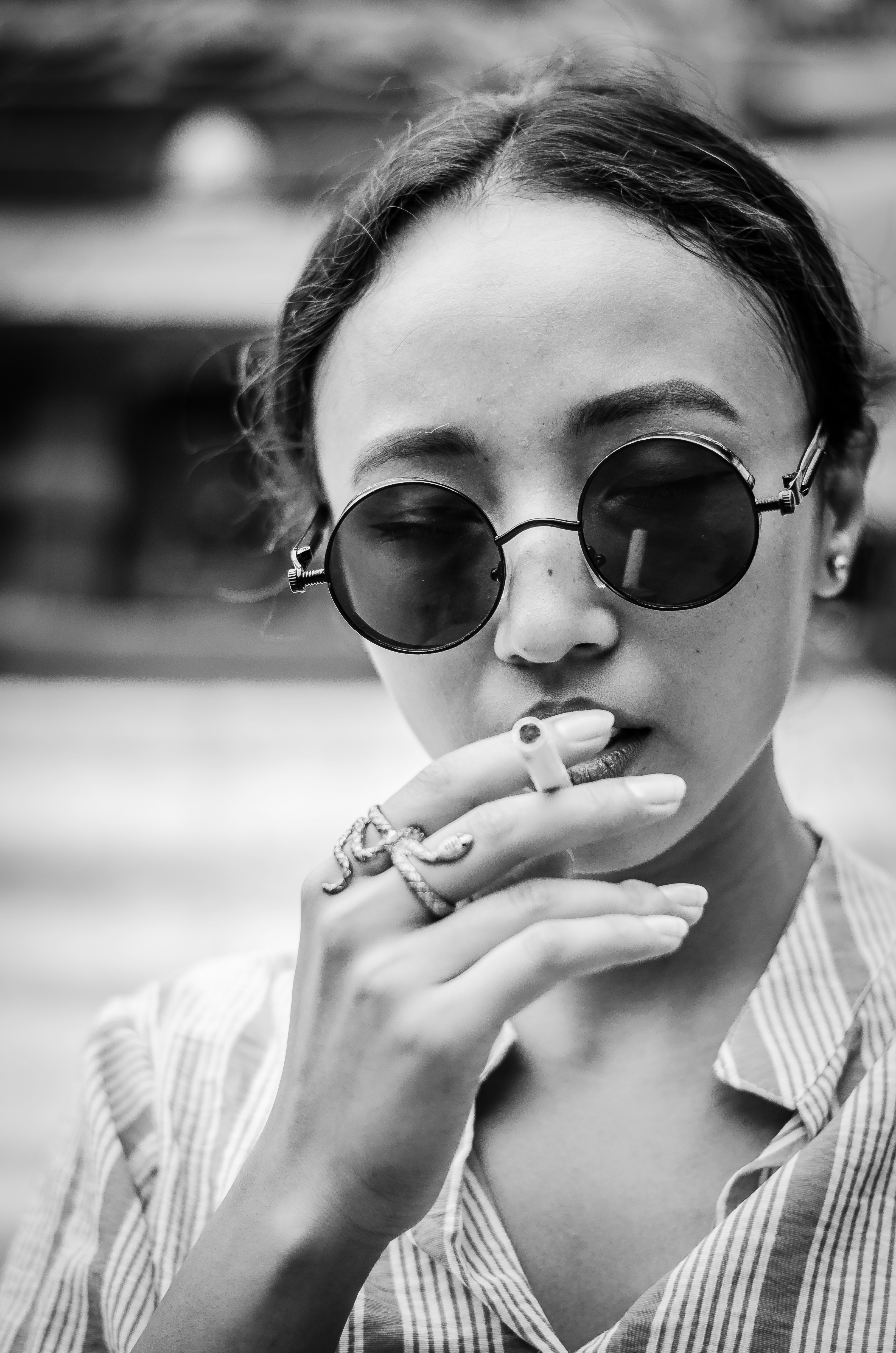Young woman in round sunglasses holds a cigarette, exuding a contemplative vibe. Her striped shirt and accessories add to the overall aesthetic.