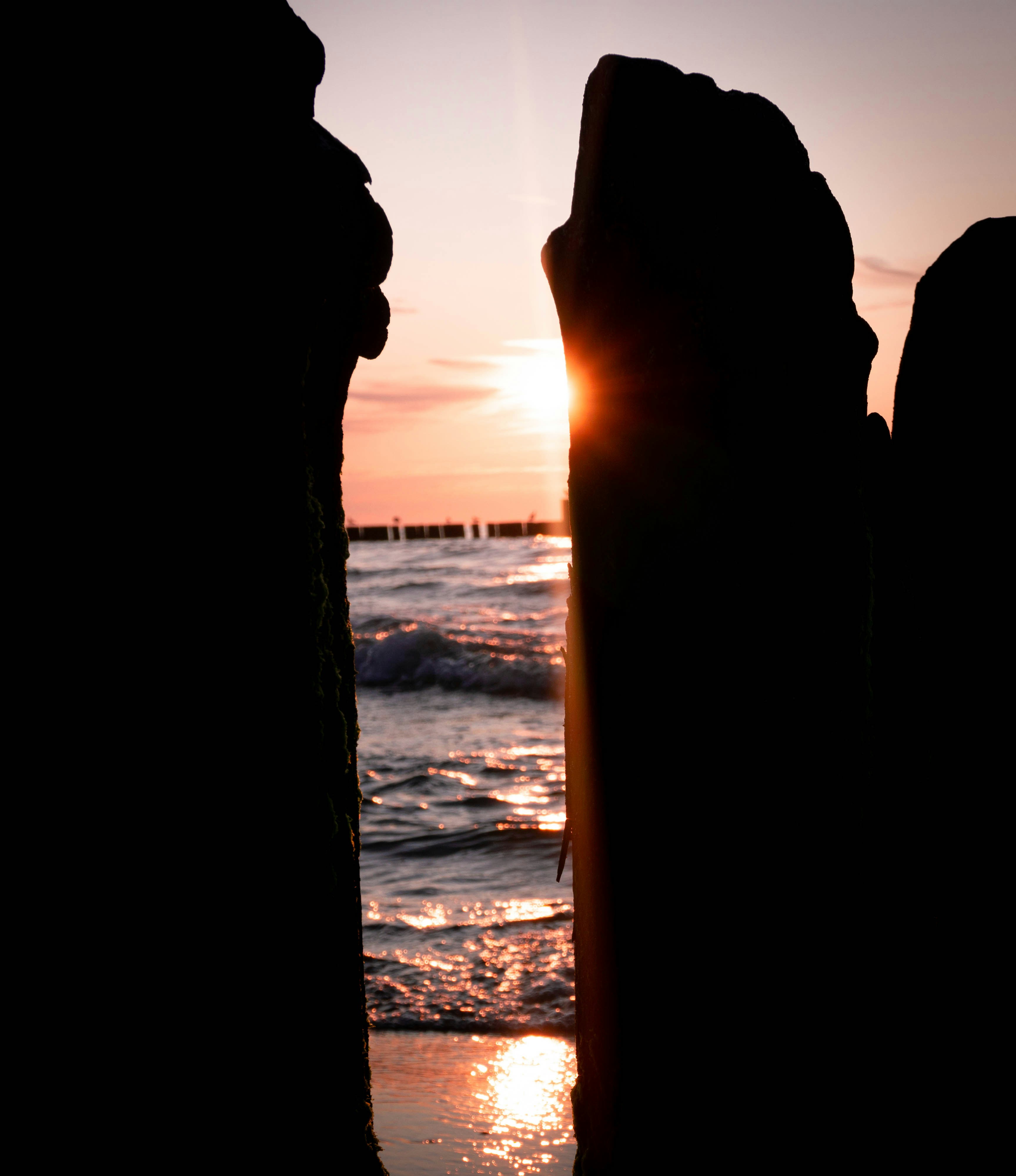 Silhouetted wooden posts frame a vibrant sunset over the ocean, casting reflections on the water's surface.