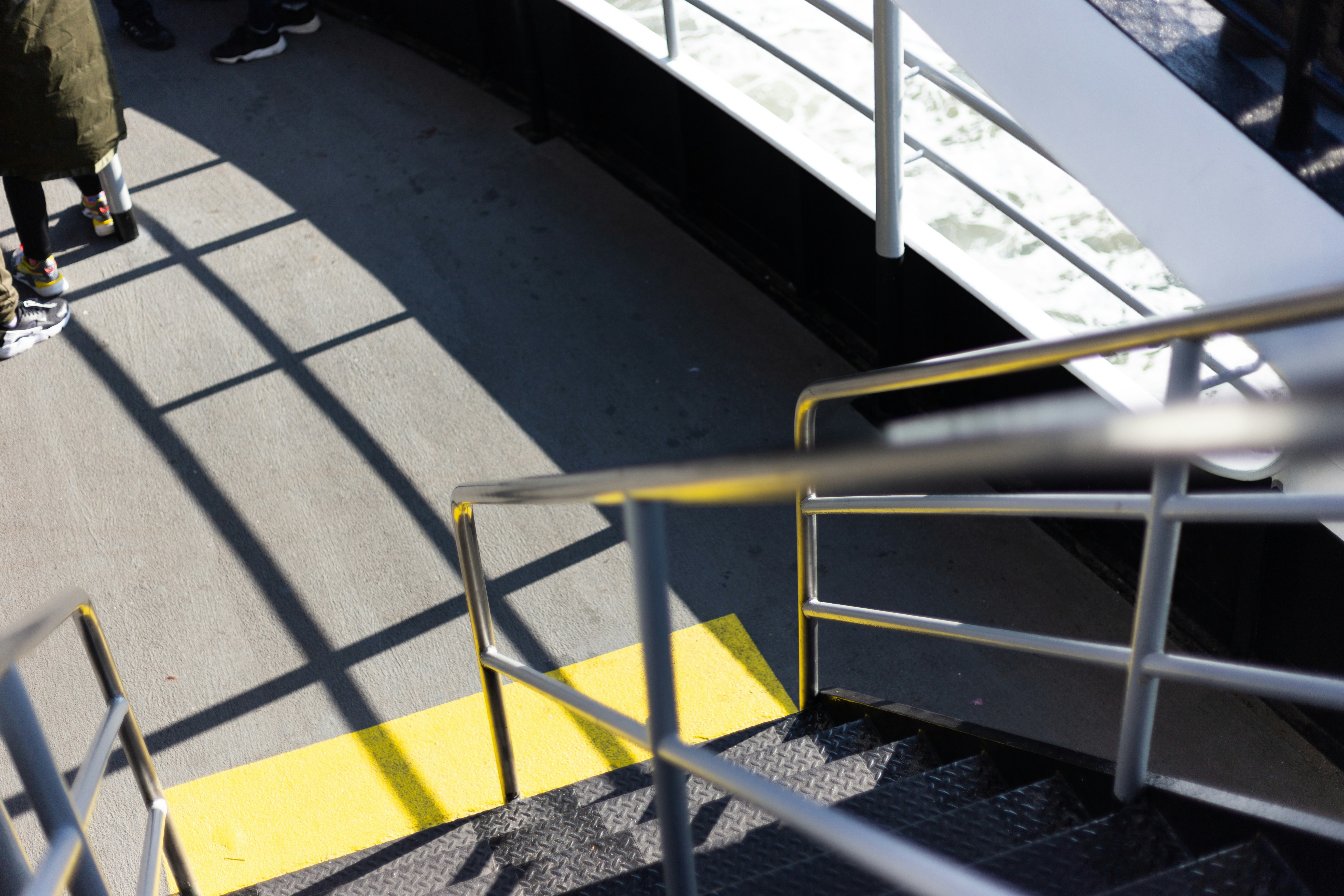 Metal staircase leading down with sunlight casting shadows on the concrete surface, featuring a bright yellow safety strip.