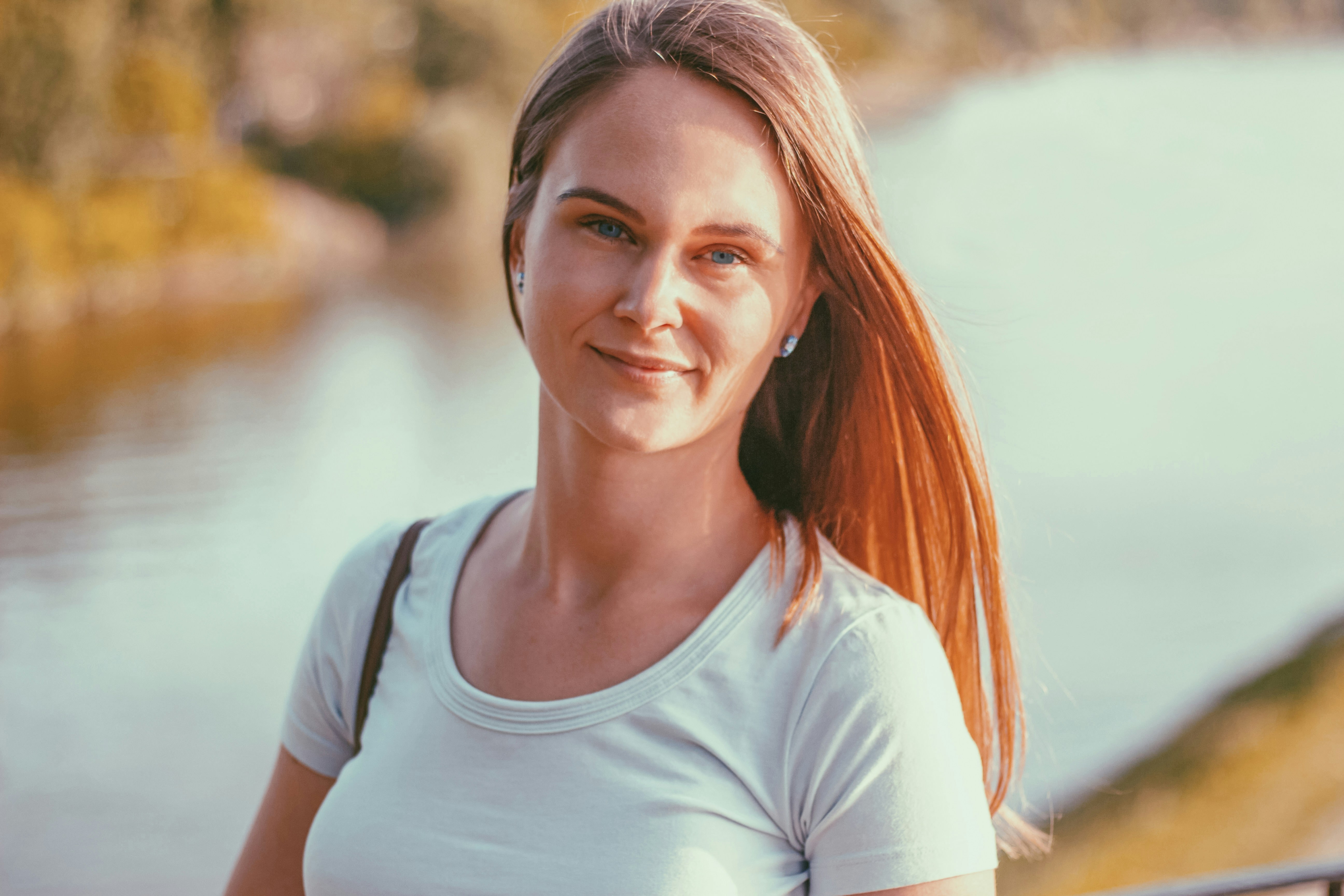 Smiling woman with flowing hair stands by a serene river under warm sunlight.
