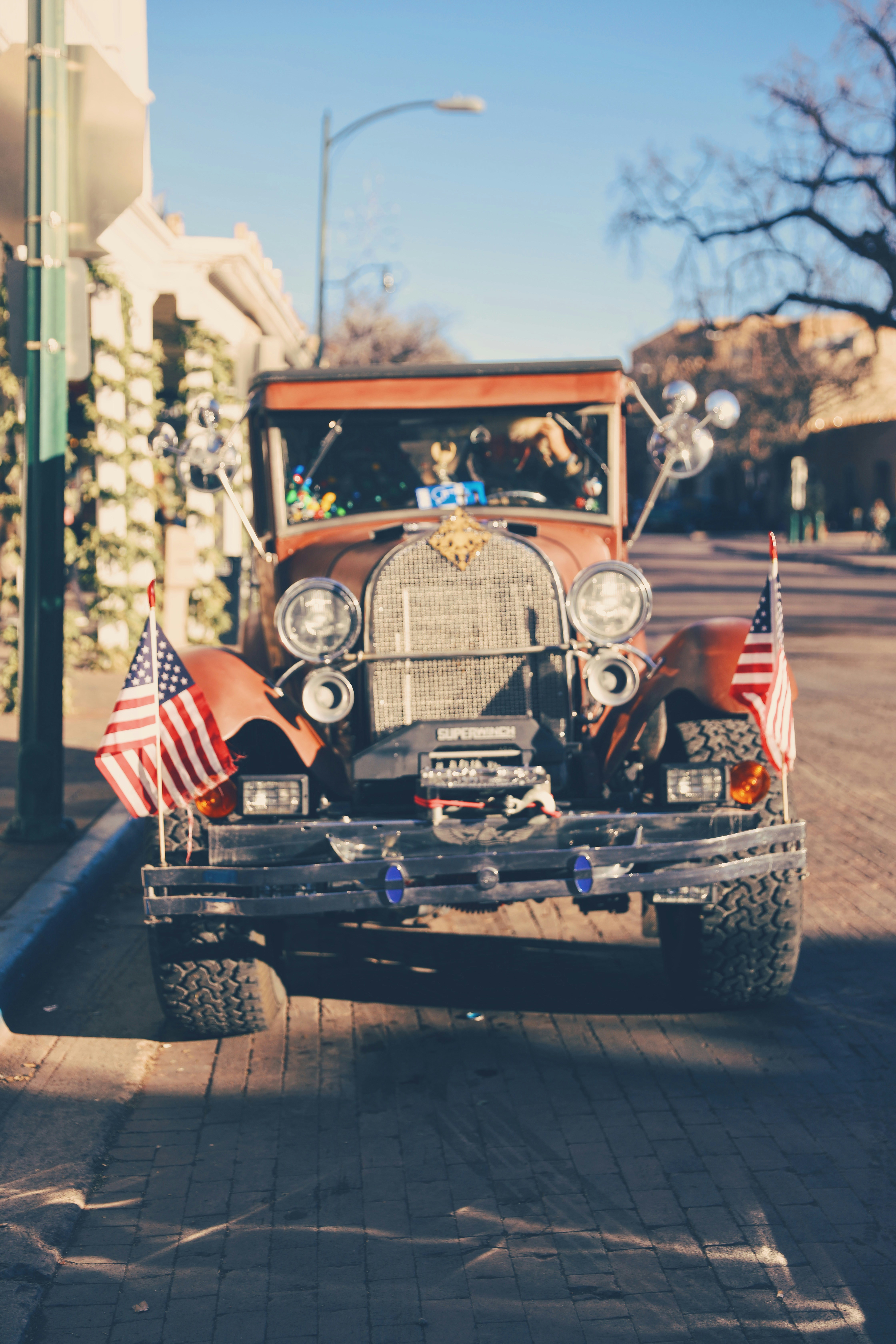 Flagged Chrome: A 1930s brass-era car parked on a sunlit street with flags fluttering at the curb
