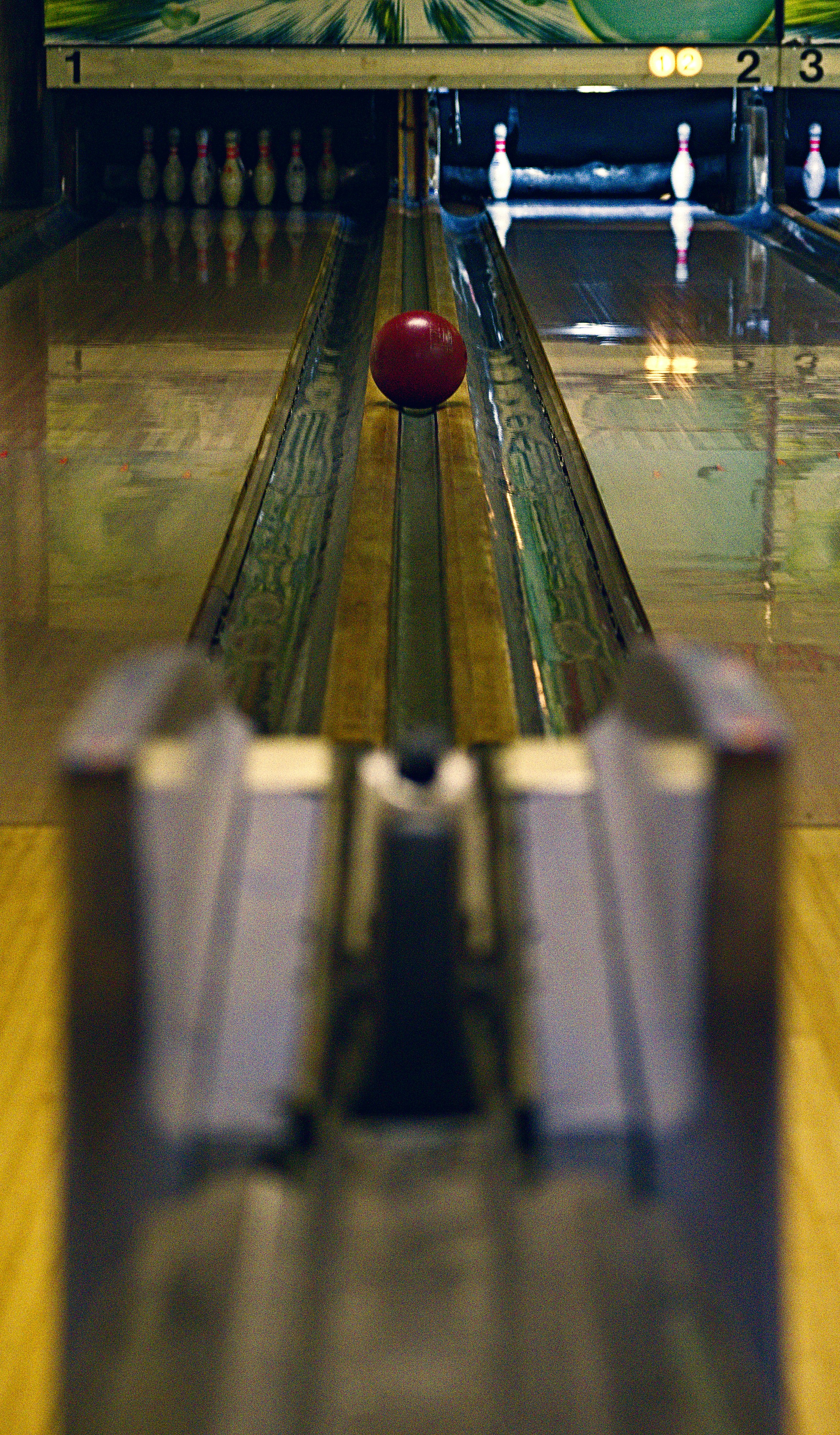 A close-up view of a bowling alley lane with a red bowling ball rolling towards the pins, framed by the bowling ball return system.
