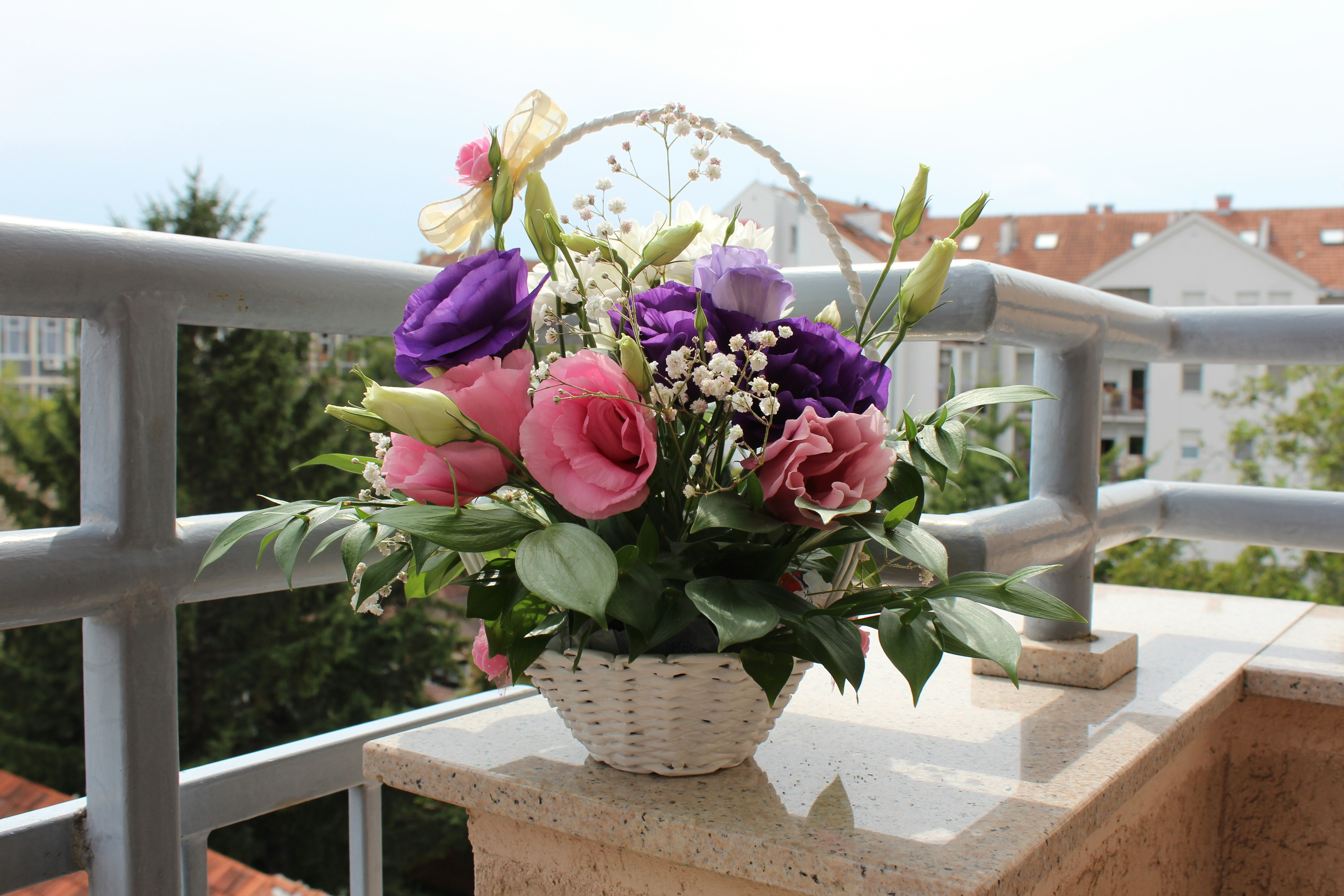 Vibrant arrangement of pink and purple flowers in a woven basket, positioned on a balcony railing with a view of buildings in the background.