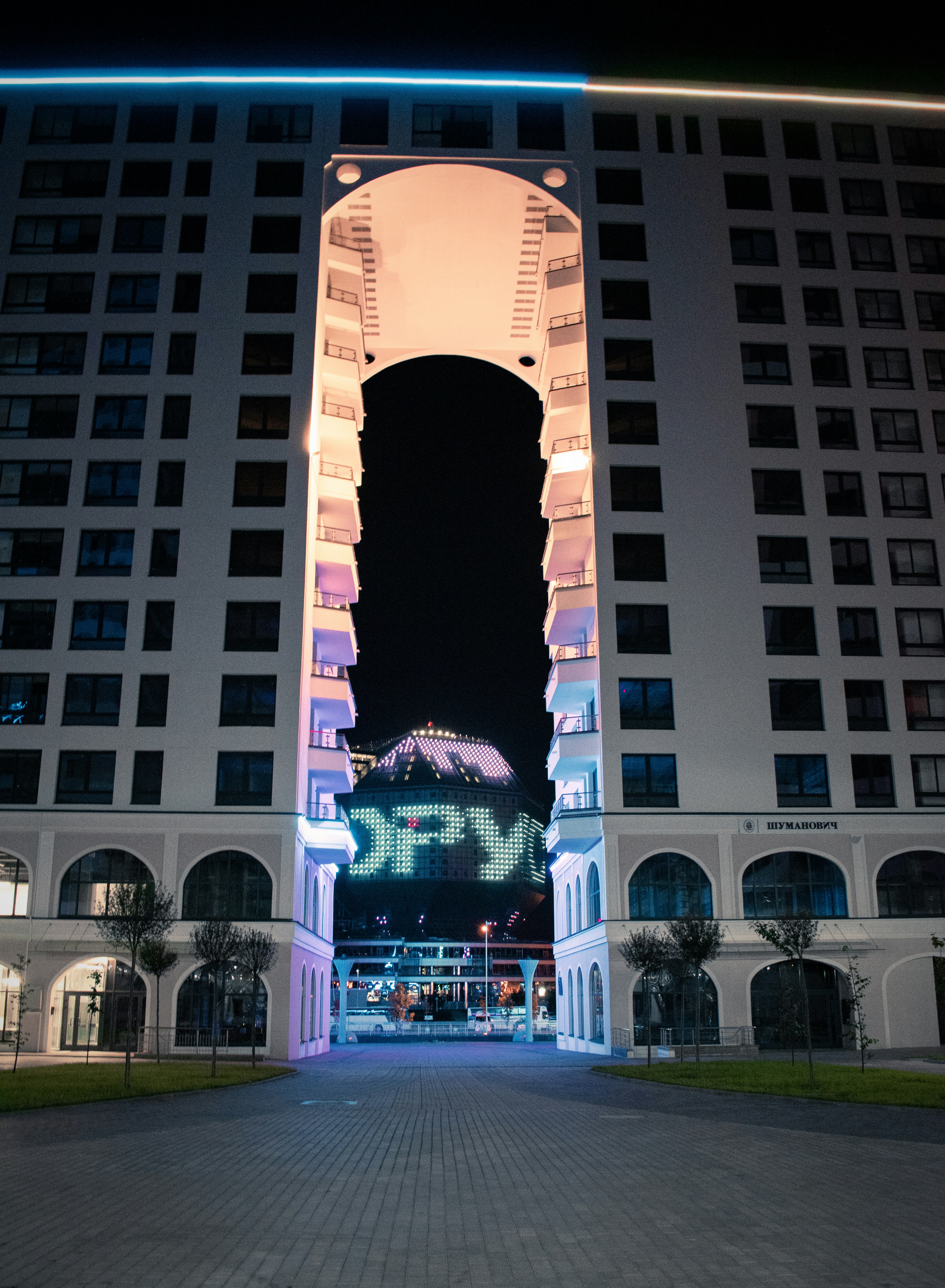 Illuminated architectural archway framed by a modern building, revealing a colorful display in the background. The scene captures the essence of urban nightlife.