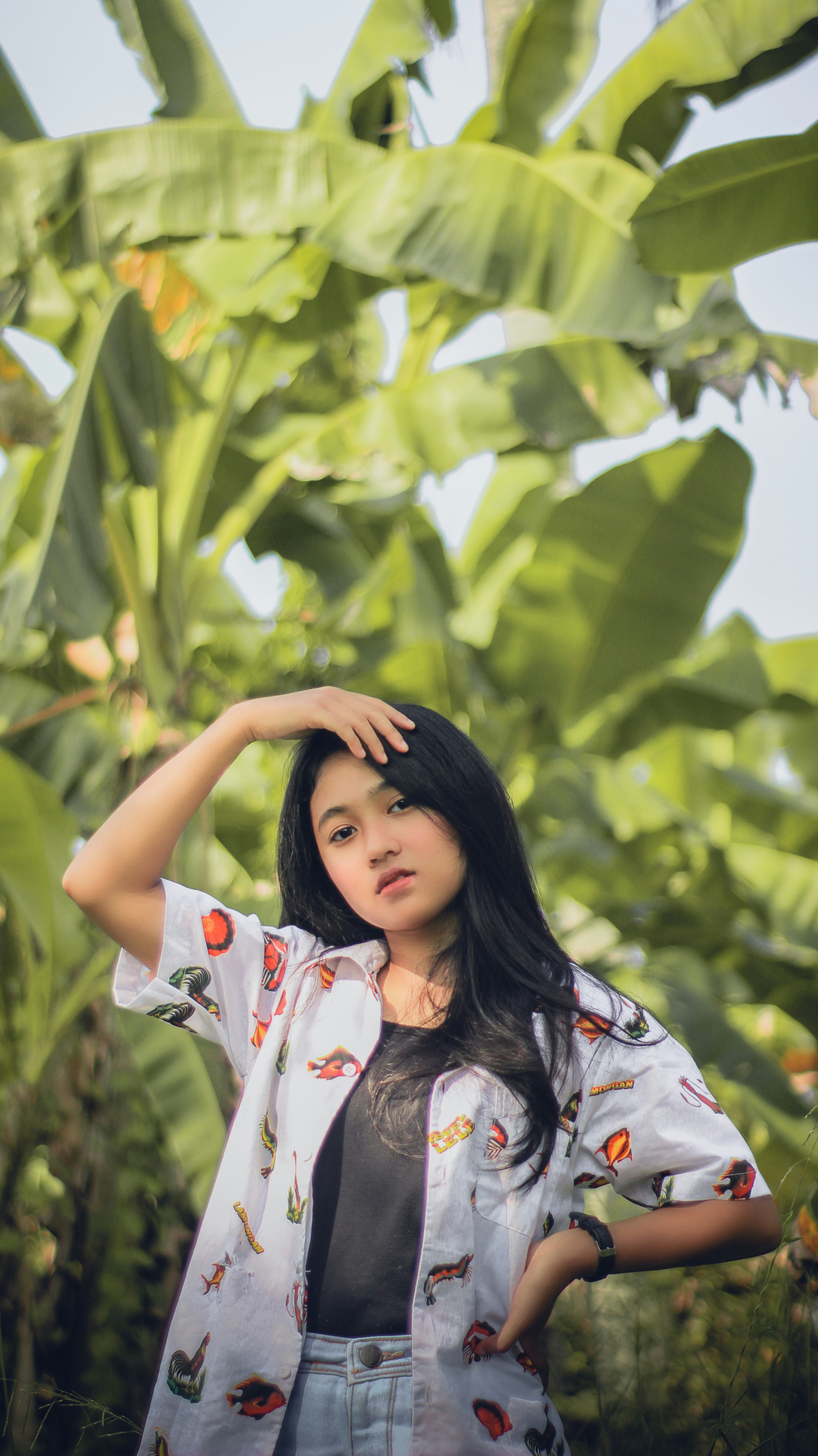 A young woman poses thoughtfully against a vibrant backdrop of banana leaves, showcasing a playful shirt design. The setting evokes a sense of tranquility and connection with nature.