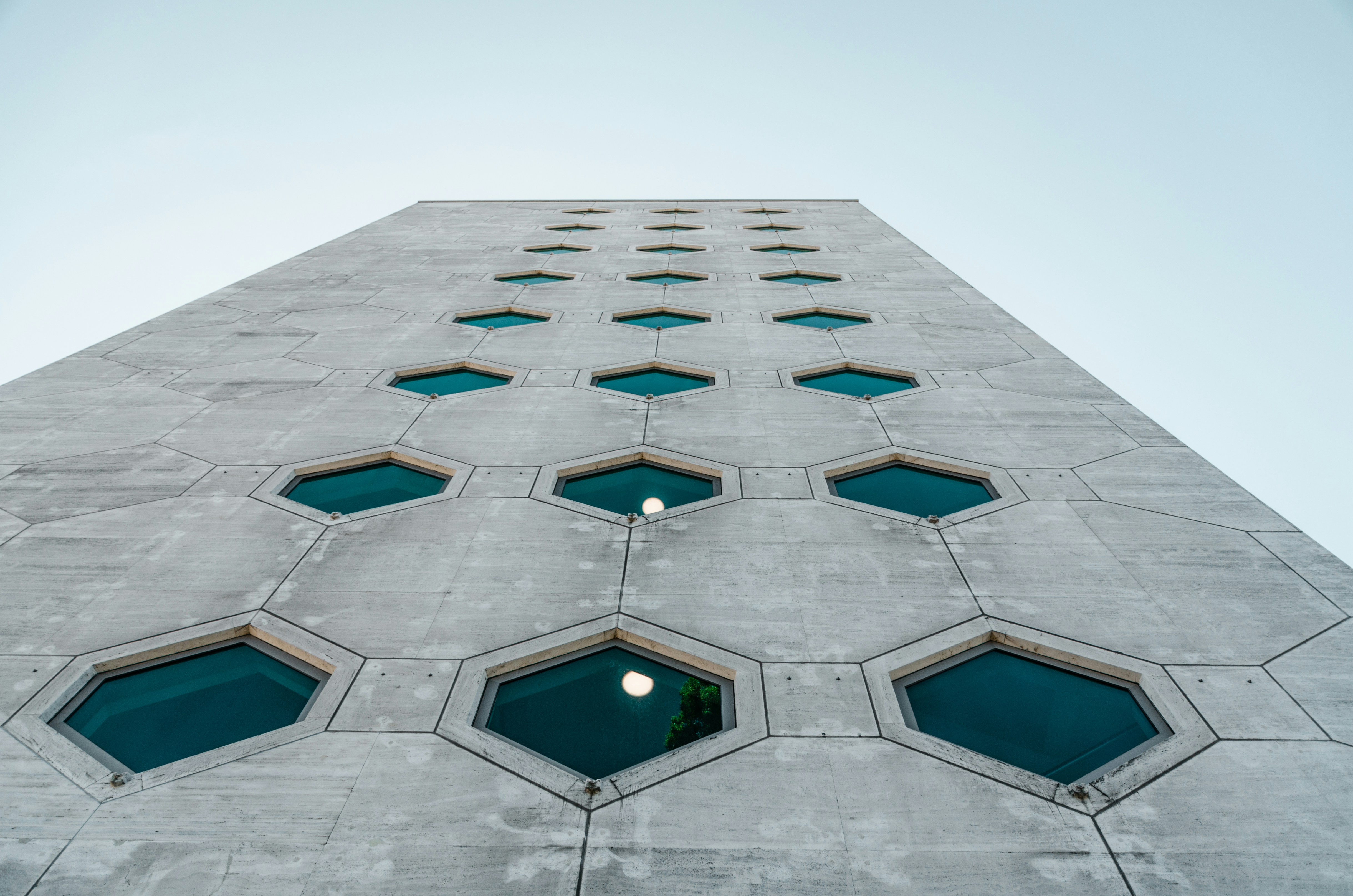 Concrete building facade featuring hexagonal windows reflecting the sky and surrounding elements.