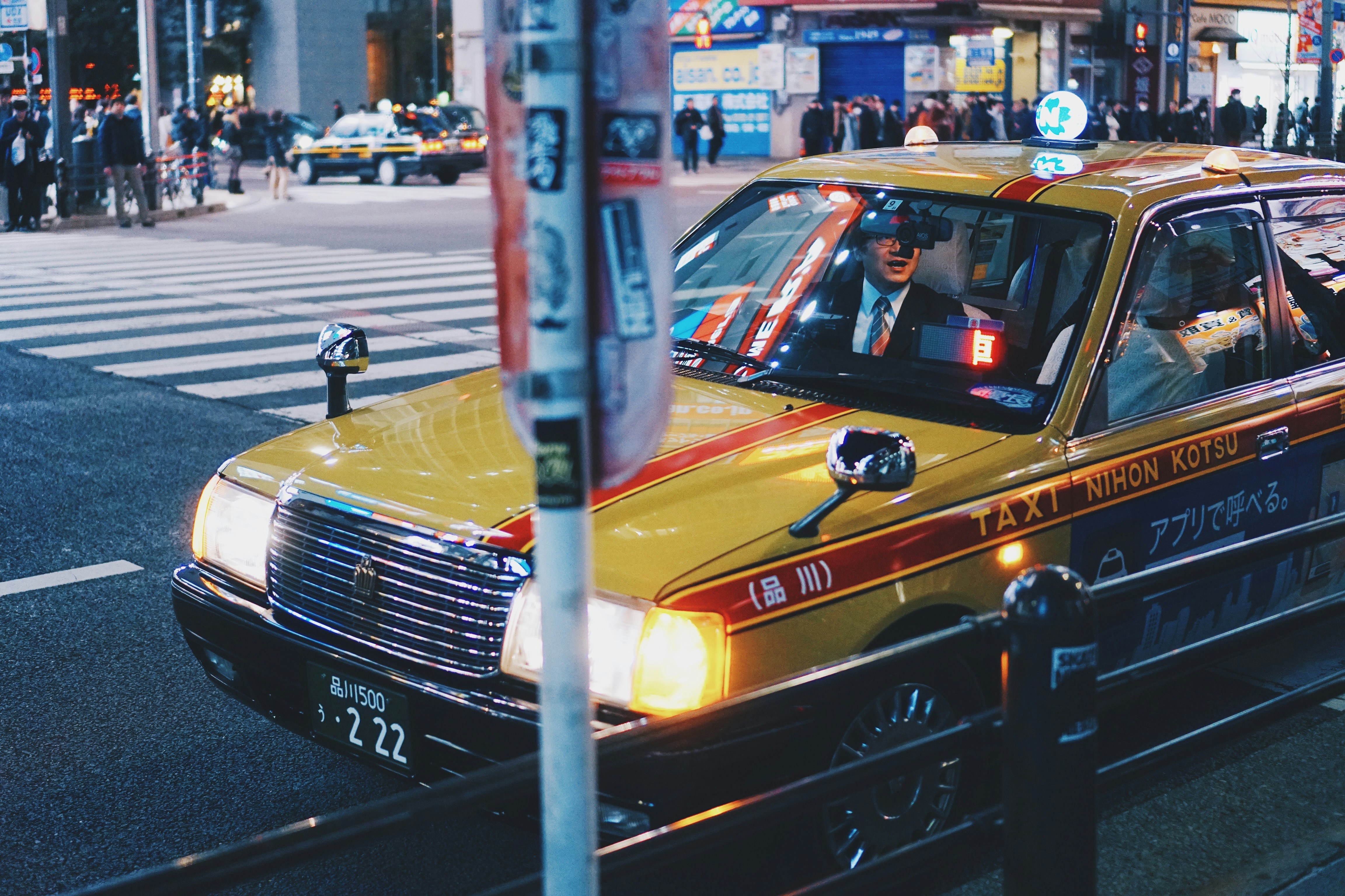 Yellow taxi navigating a bustling city intersection, illuminated by vibrant neon lights and surrounded by pedestrians. The driver's focused expression adds to the urban atmosphere.