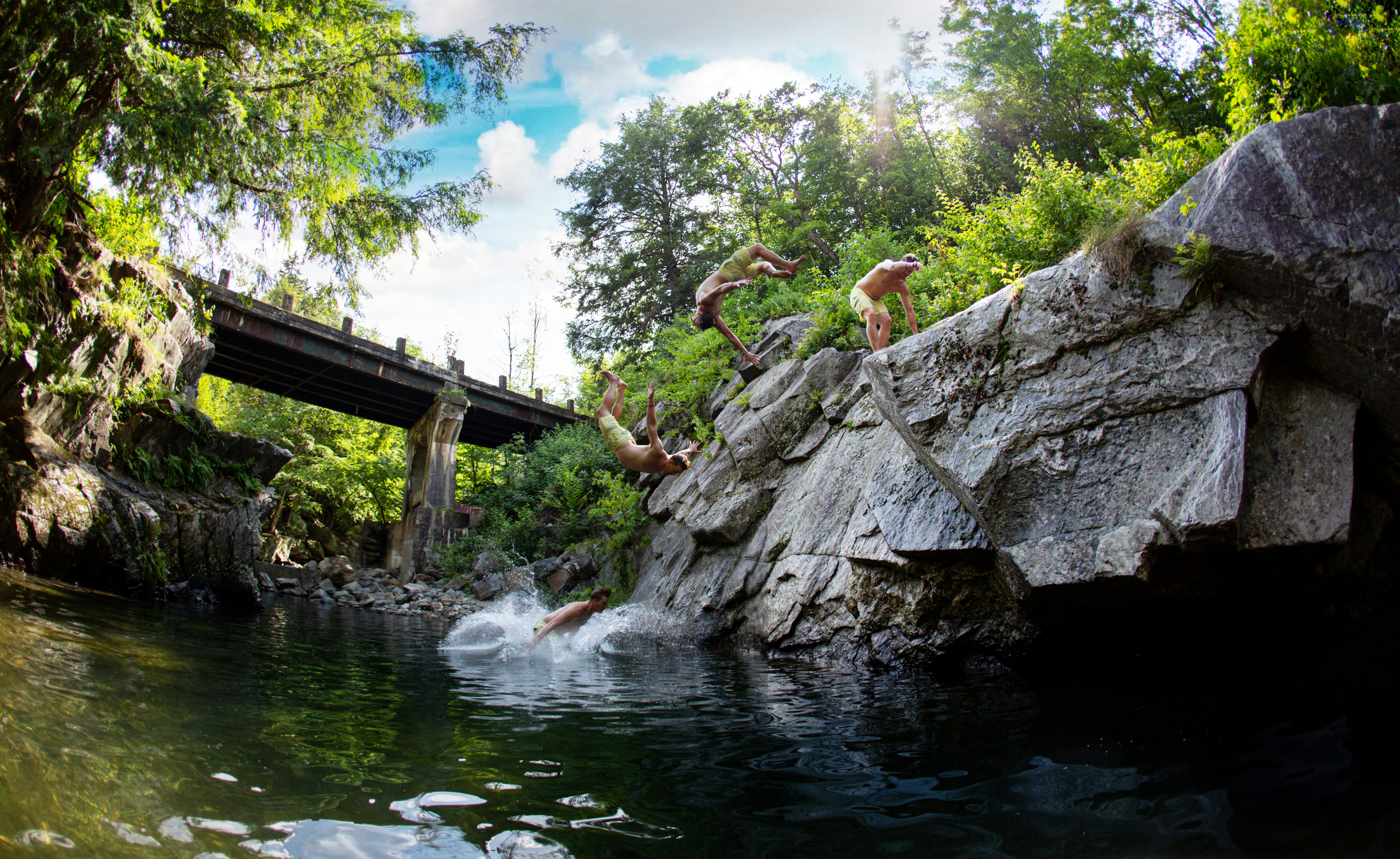 Four men about to jump on lake surrounded with tall and green trees ...