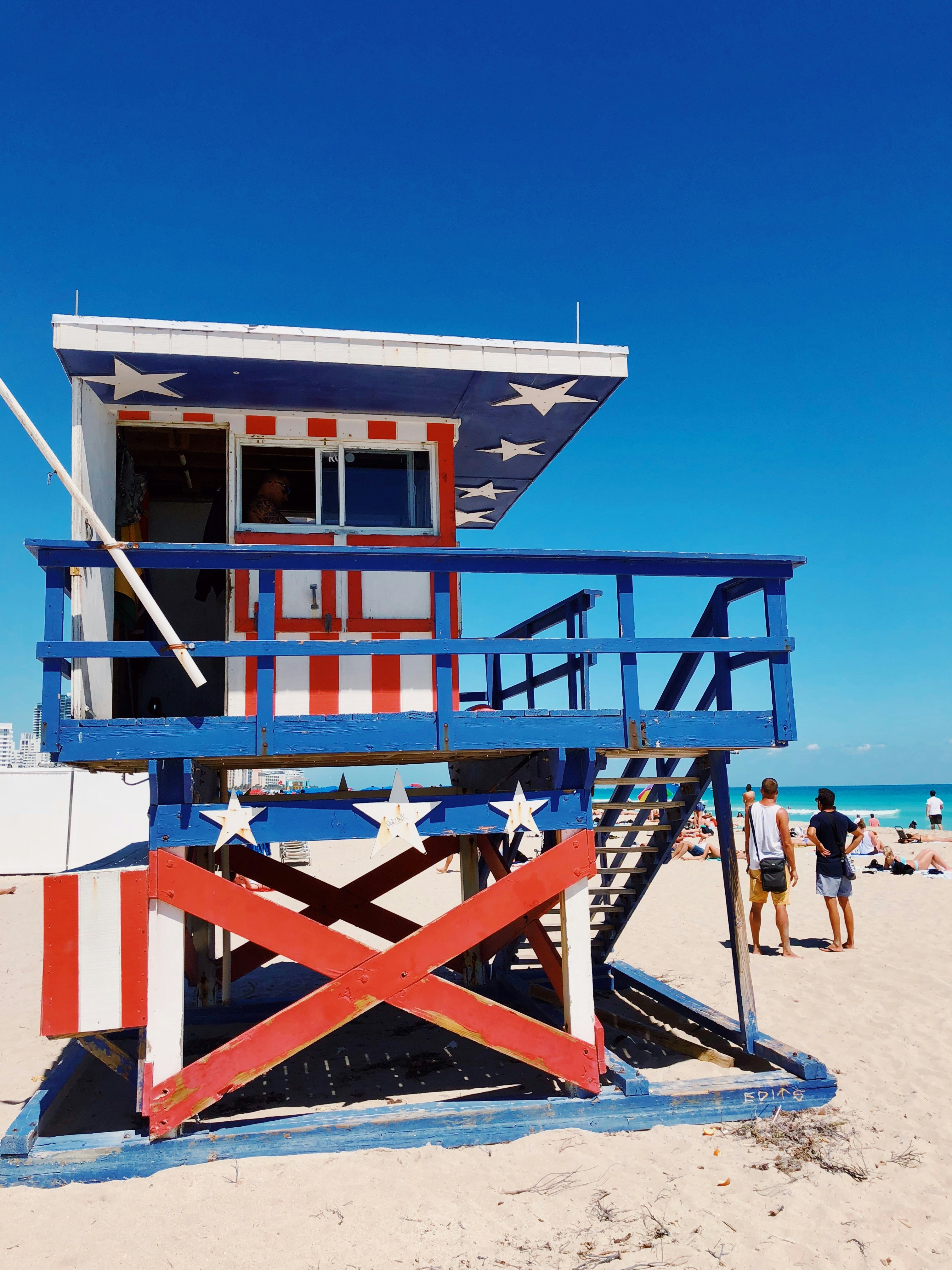 Colorful lifeguard stand adorned with red, white, and blue stripes overlooking a sandy beach filled with sunbathers.