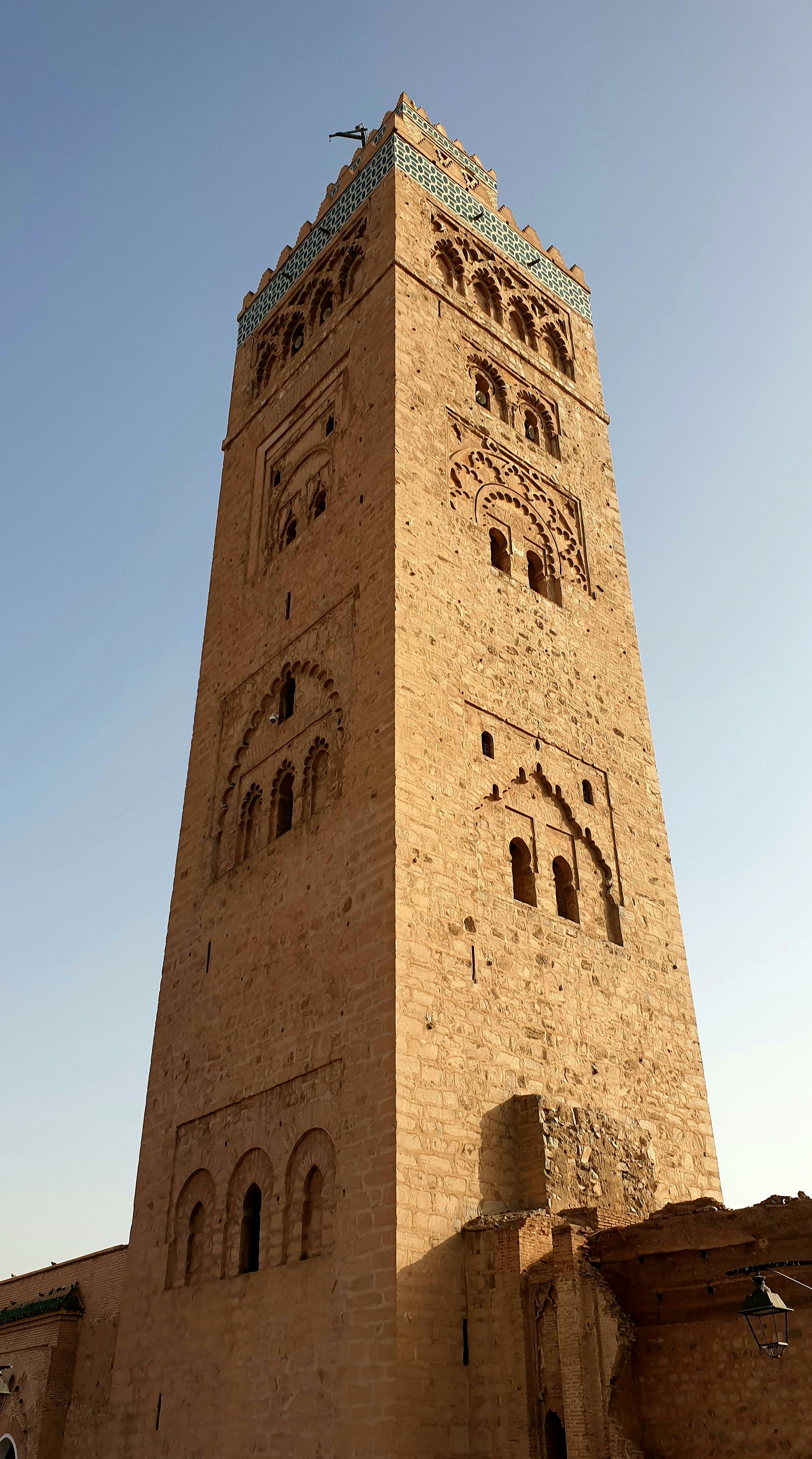 A tall, ancient stone minaret rises against a clear blue sky, its ornate arches and turquoise-tiled cornice detailing the structure.