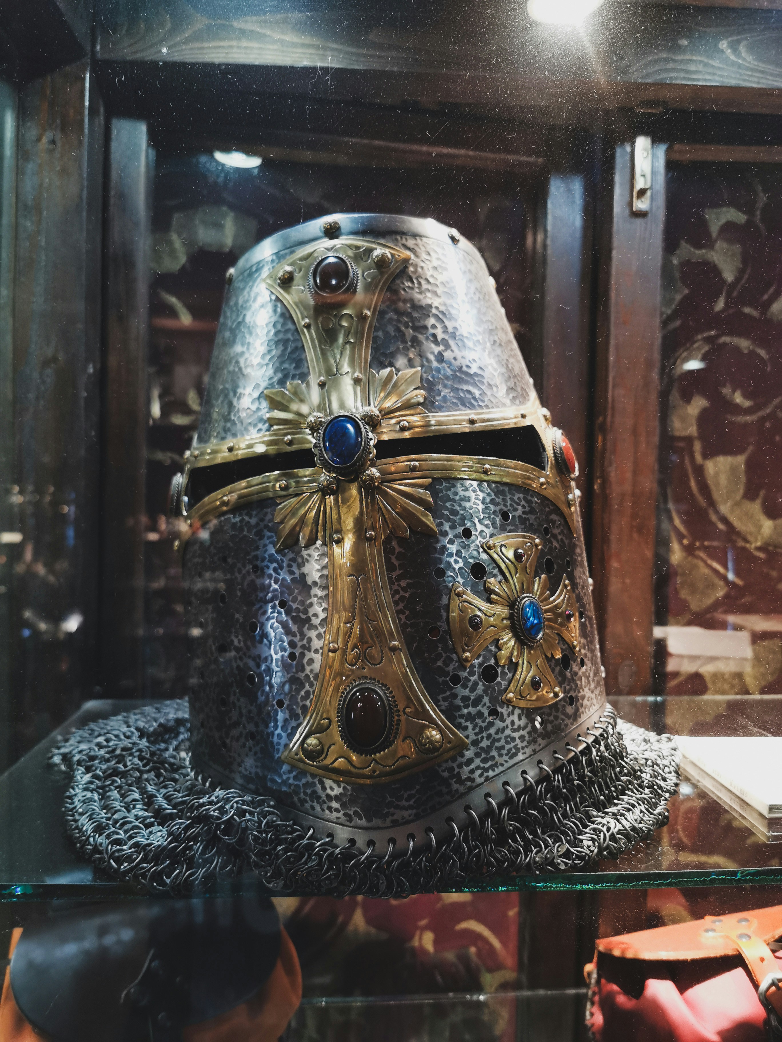 Intricately designed medieval helmet adorned with gold accents and colorful gemstones, displayed in a glass case.