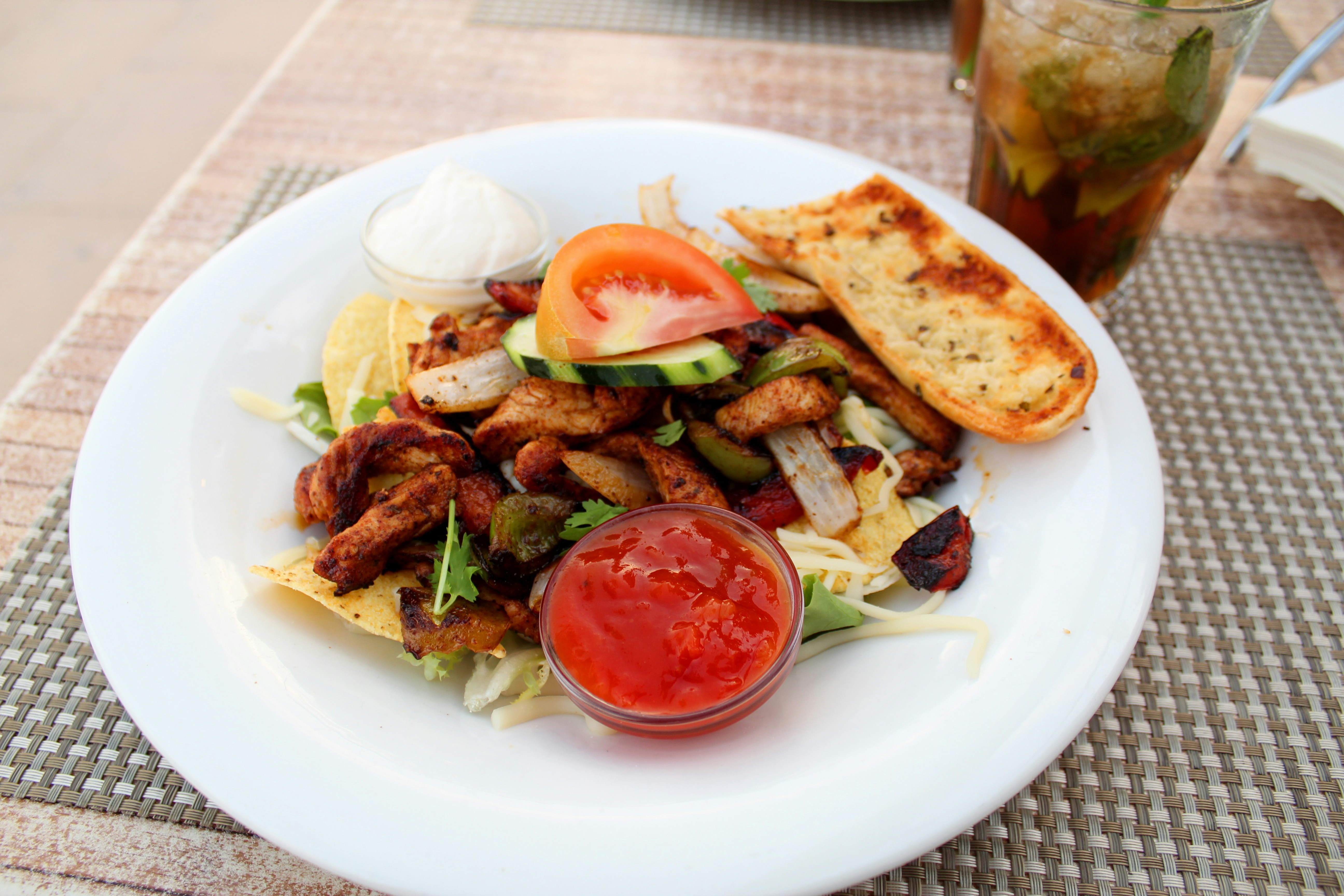 A vibrant plate of nachos topped with grilled meat, fresh vegetables, and sauces, accompanied by a toasted bread slice and a refreshing drink.
