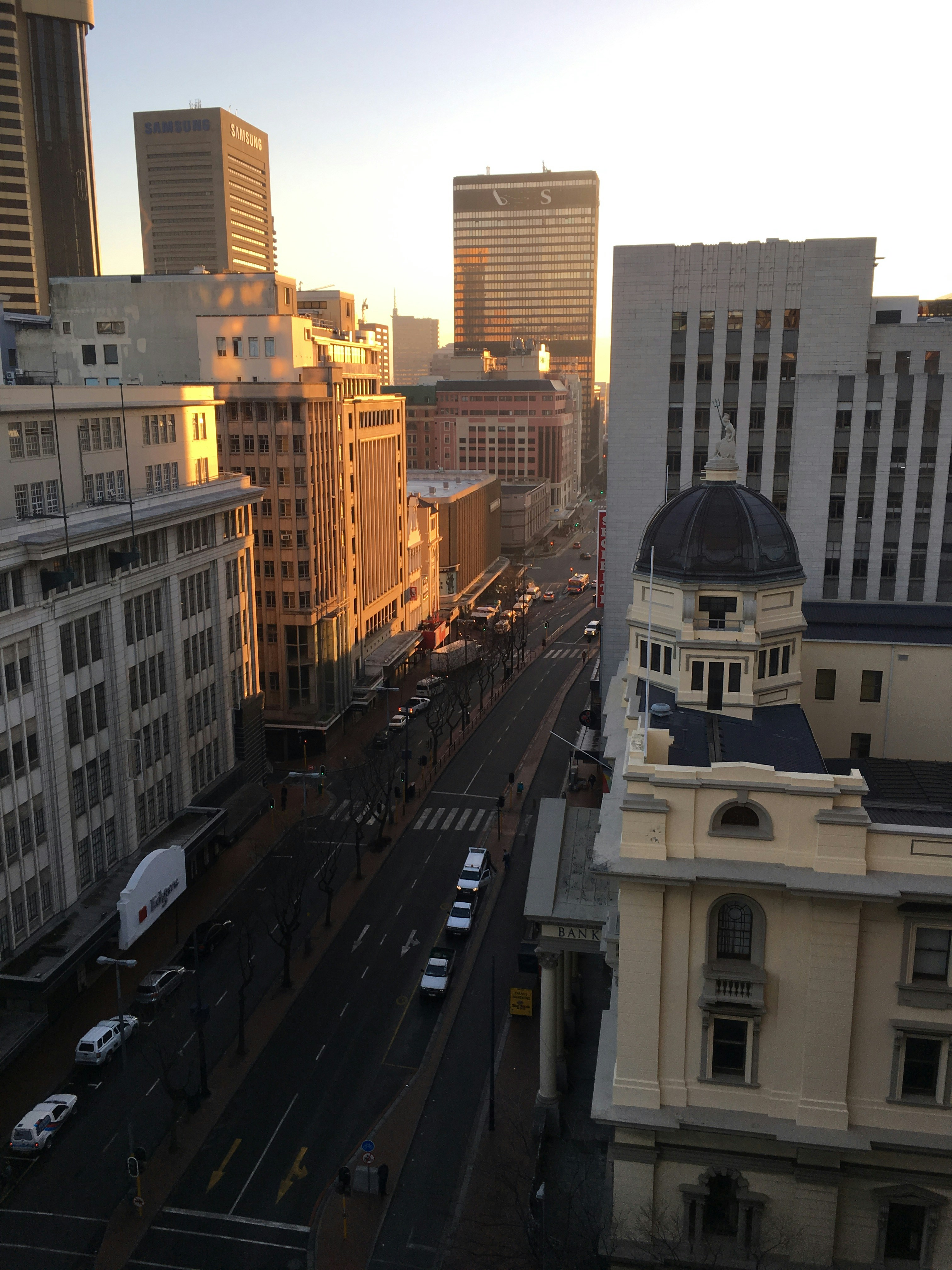 Golden sunlight casts a warm glow over a bustling city street, highlighting the architectural contrasts between historic and modern buildings.