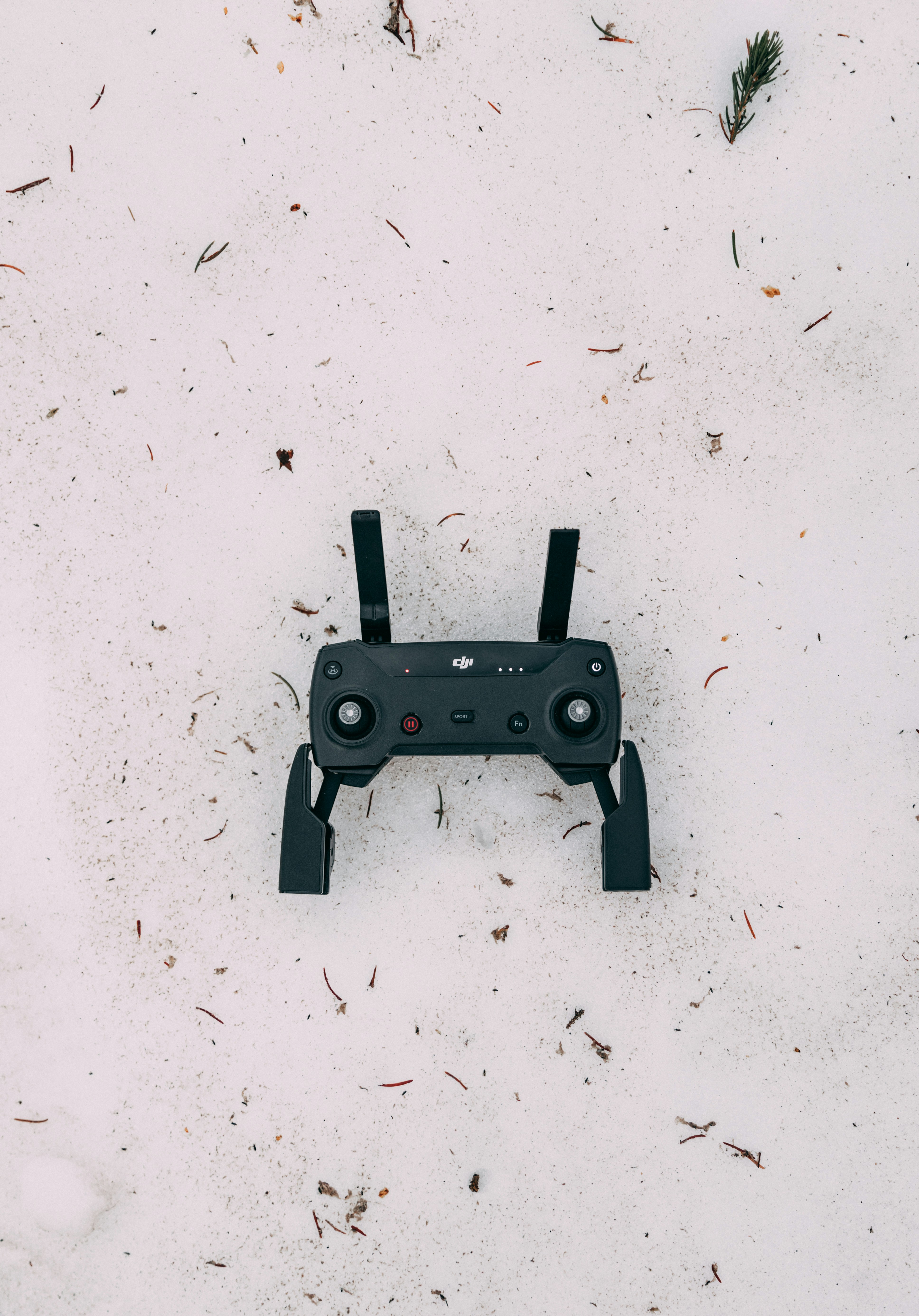 Drone controller resting on a snowy surface with scattered pine needles around it.