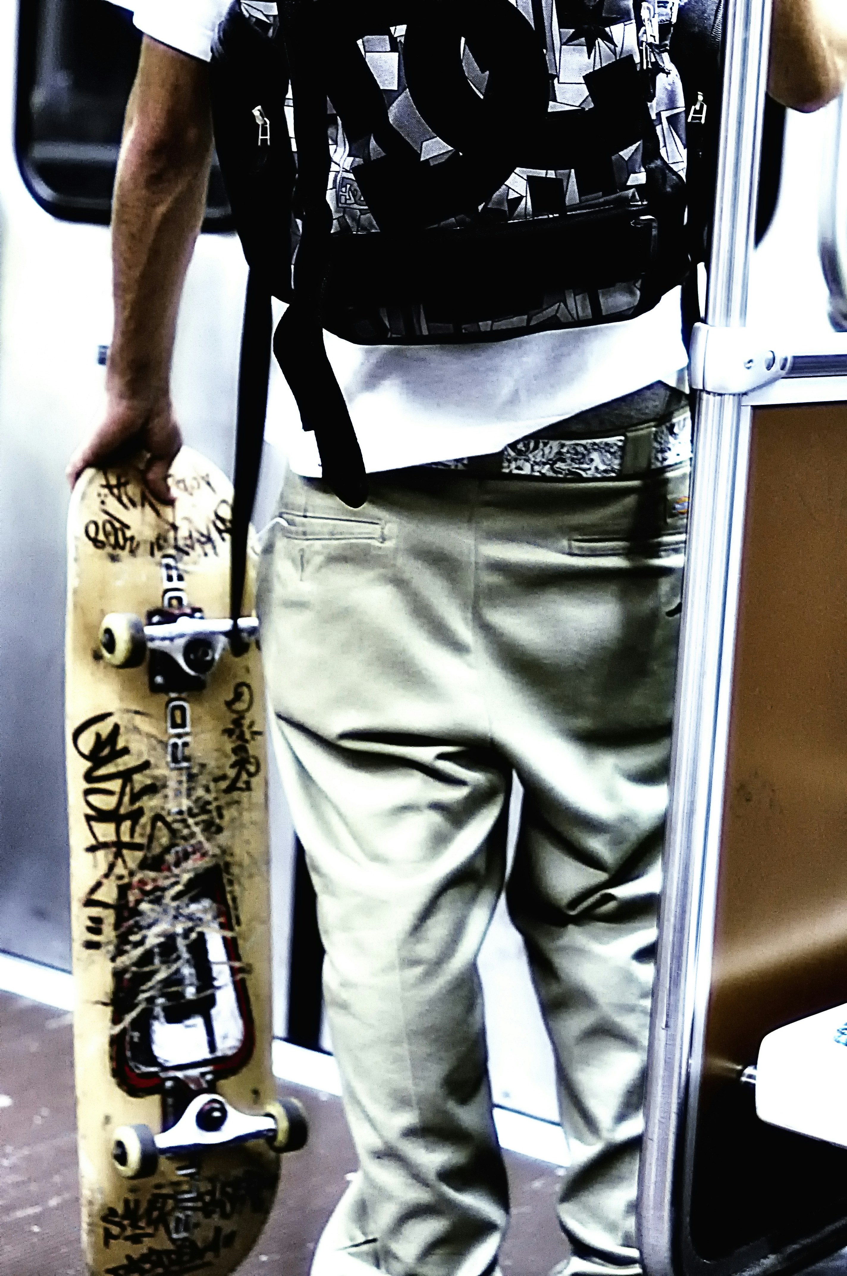 A skateboarder stands in a subway car, gripping a graffiti-adorned skateboard while wearing casual attire and a backpack. The scene captures the intersection of urban transit and skate culture.