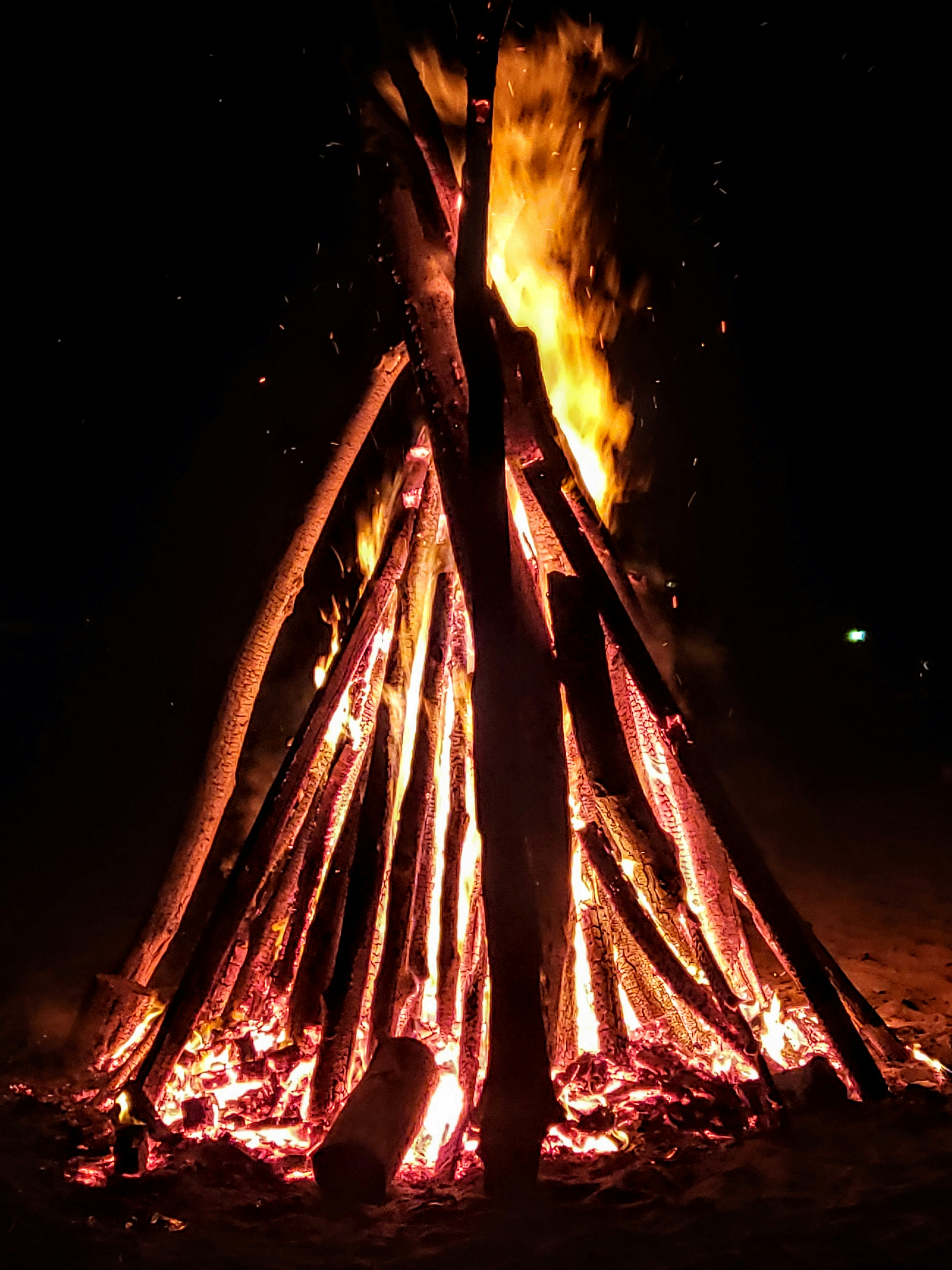 A towering bonfire crackles with vibrant flames and glowing embers against a dark backdrop, illuminating the surrounding area with warmth.