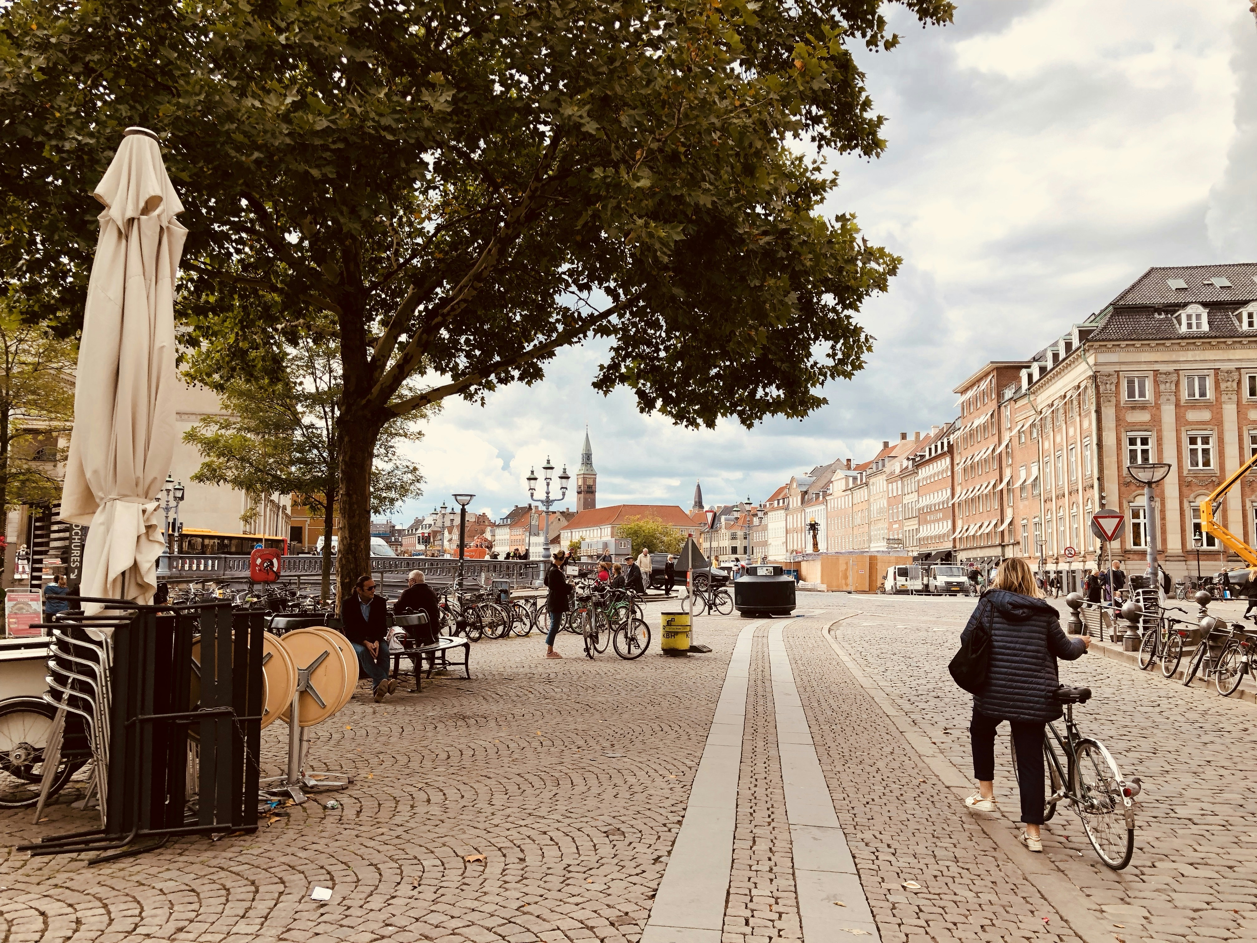 A bustling street scene featuring cyclists and pedestrians amidst charming architecture and lush trees. The atmosphere reflects a vibrant urban lifestyle.