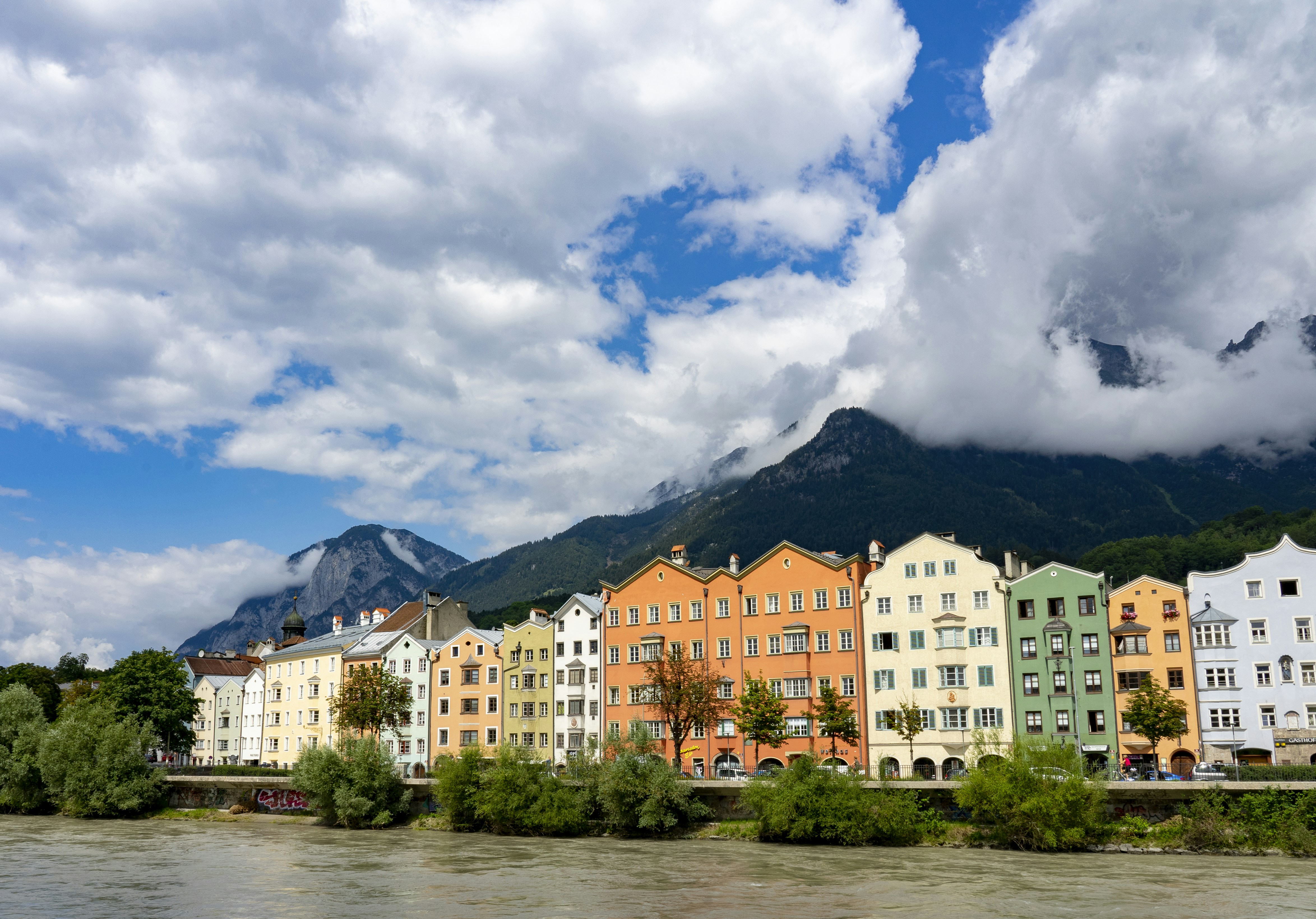 Row of vibrant houses beside a river, set against a dramatic mountainous backdrop under a partly cloudy sky.