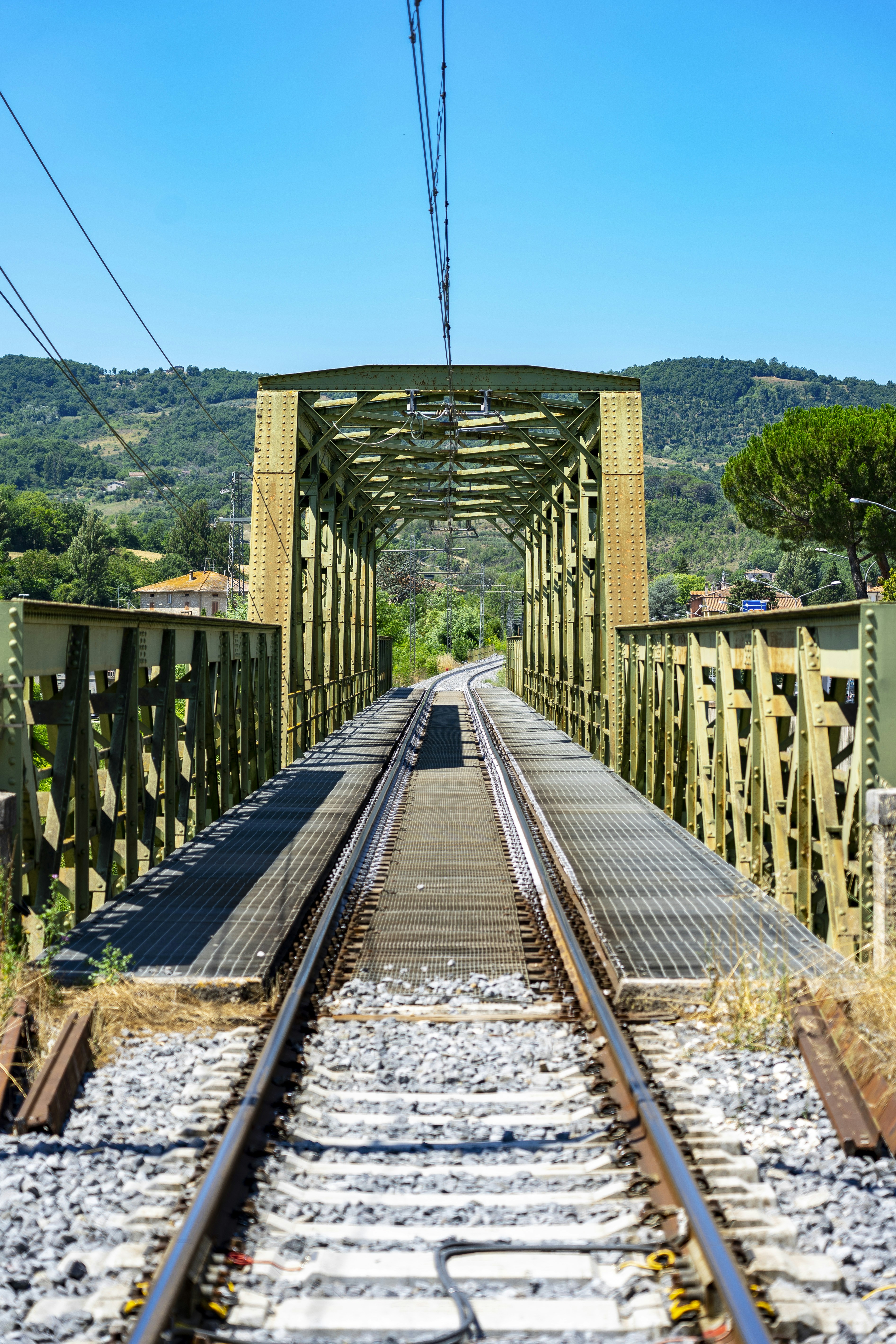 Railway tracks converge beneath a vintage metal bridge, framed by lush greenery and distant hills. A testament to engineering and nature's harmony.