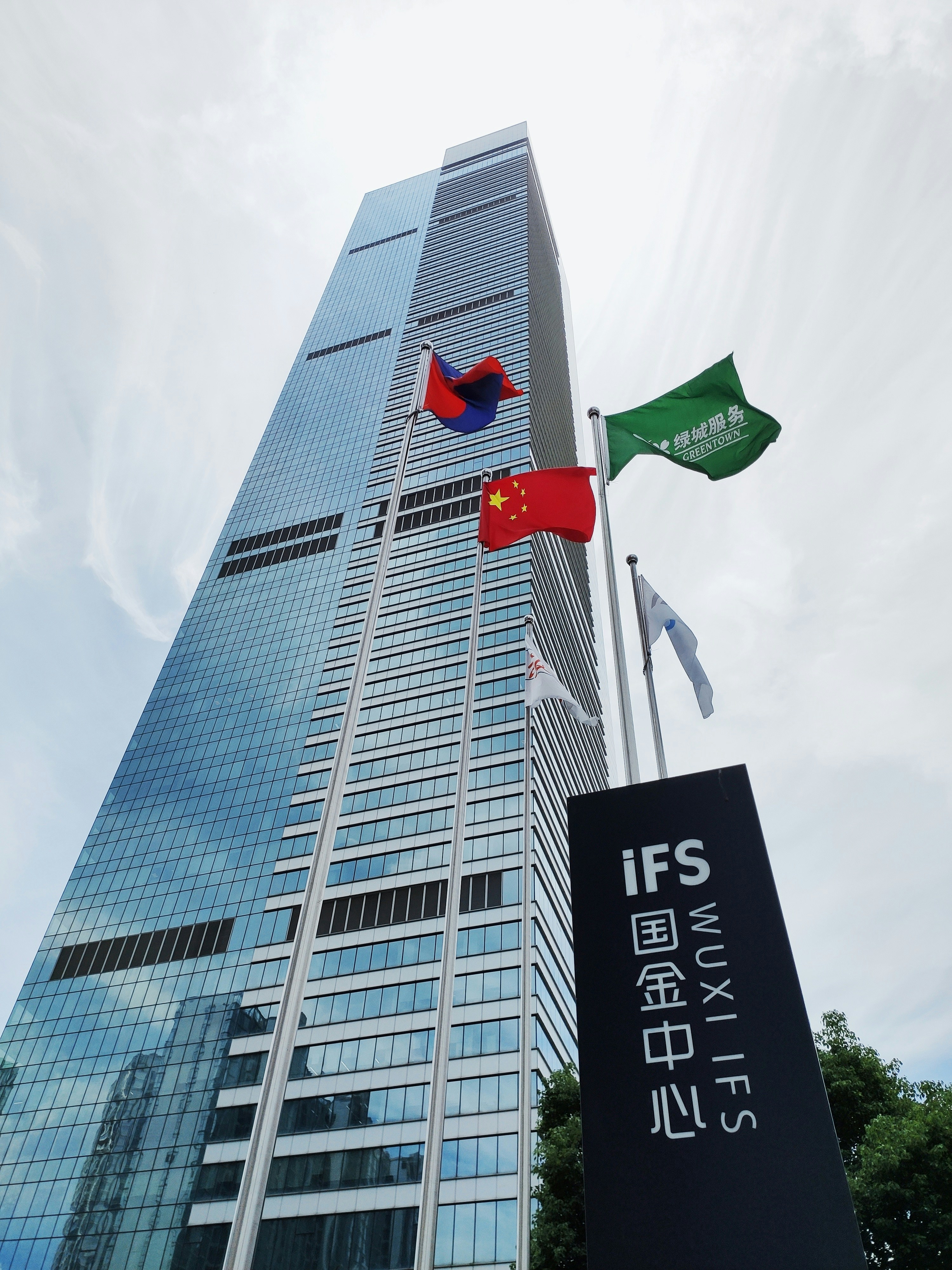 Tall glass skyscraper with multiple flags fluttering near its base against a bright blue sky.