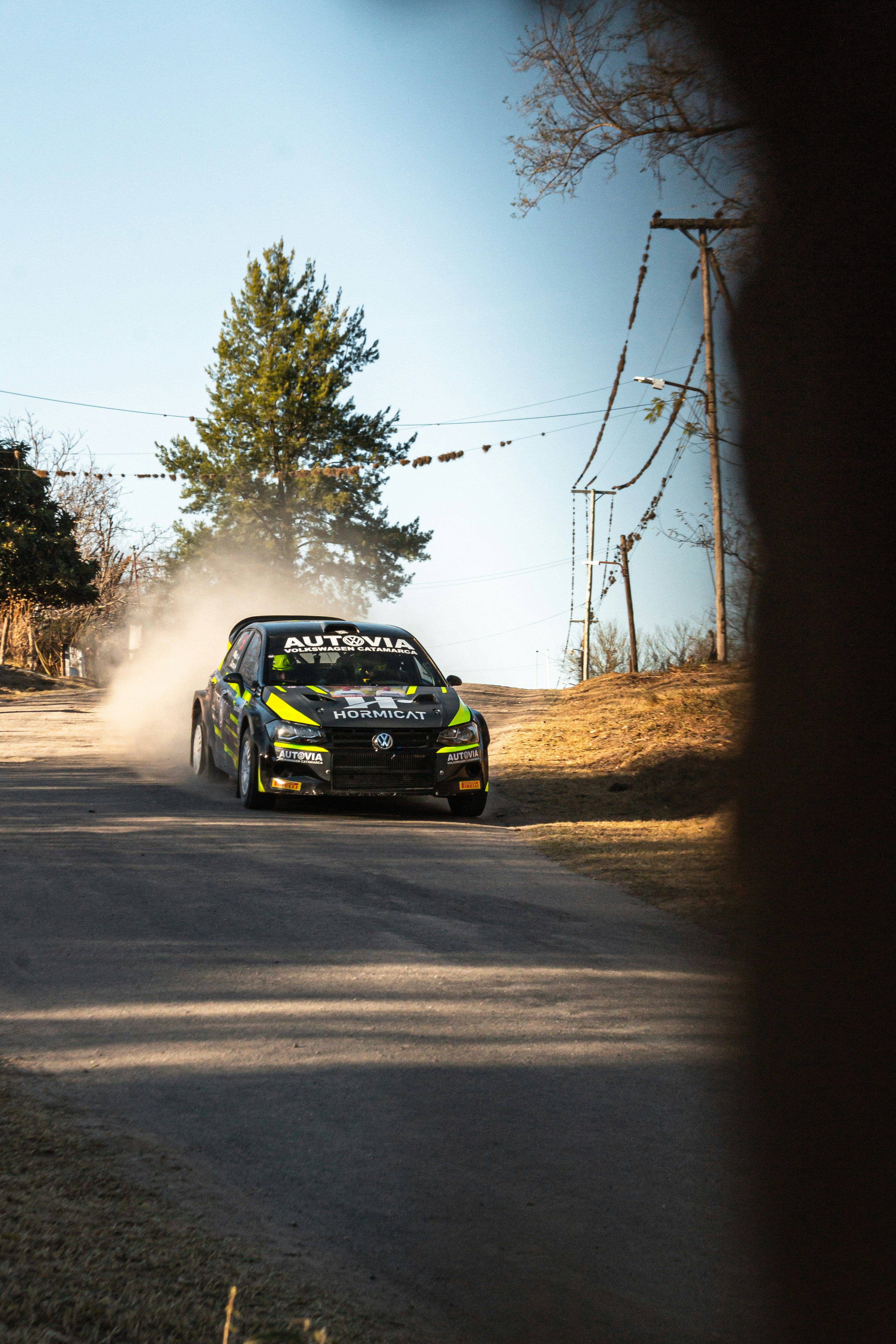 Black rally car speeding on a rural road, kicking up dust near power lines.
