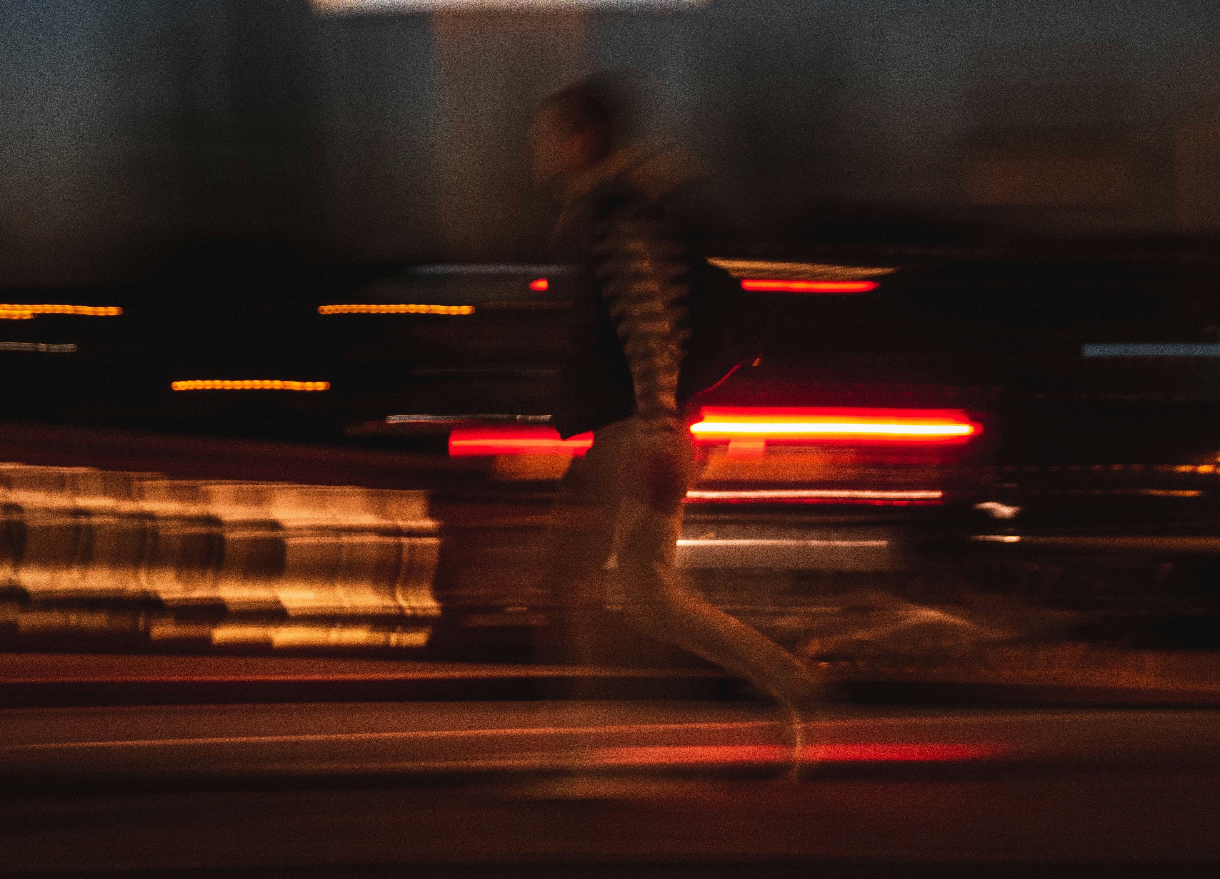 Blurry figure walking across a city street at night with streaks of car lights in the background.