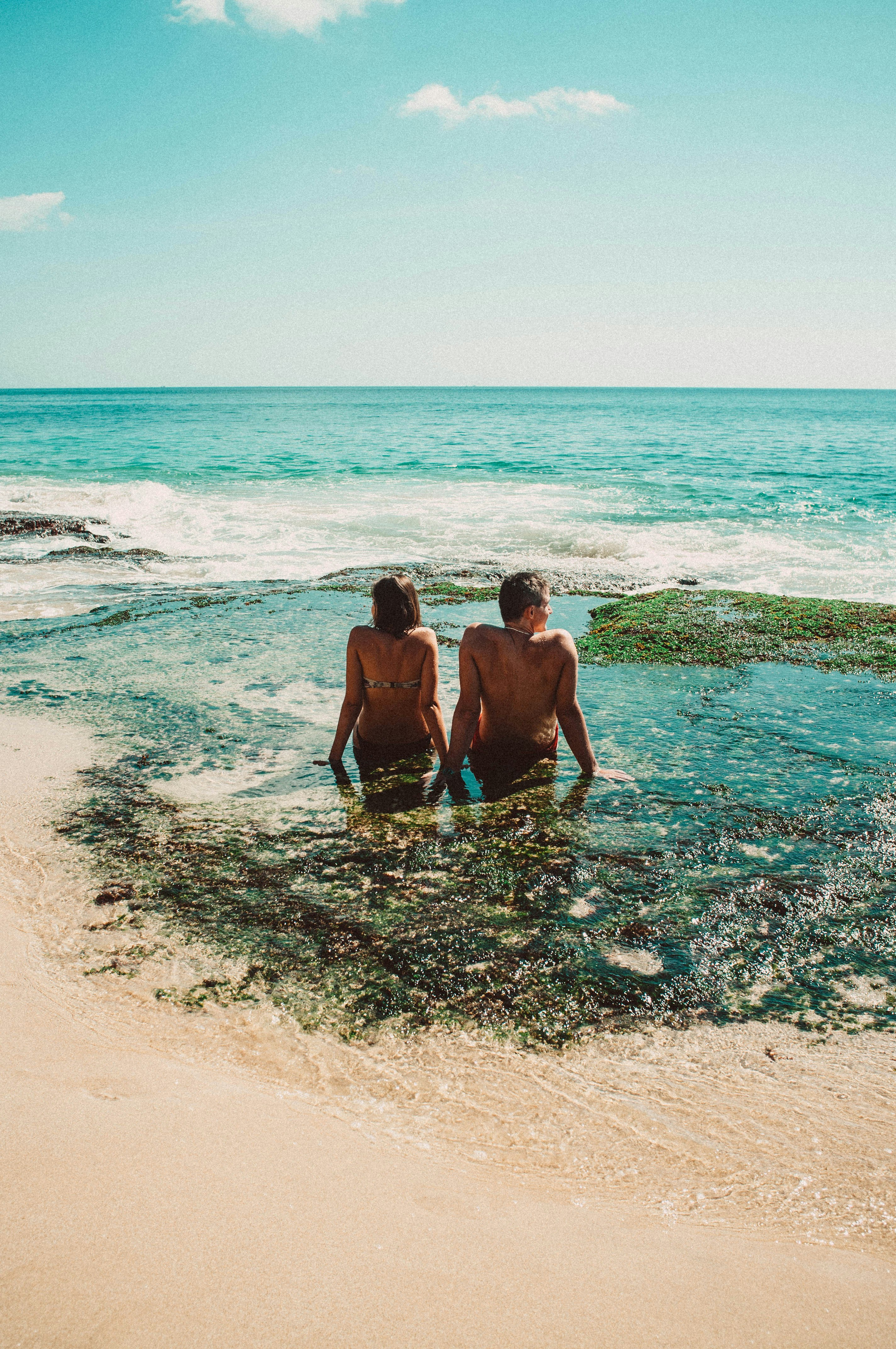 Two people sit in shallow tide pools facing the turquoise ocean under a clear sky.