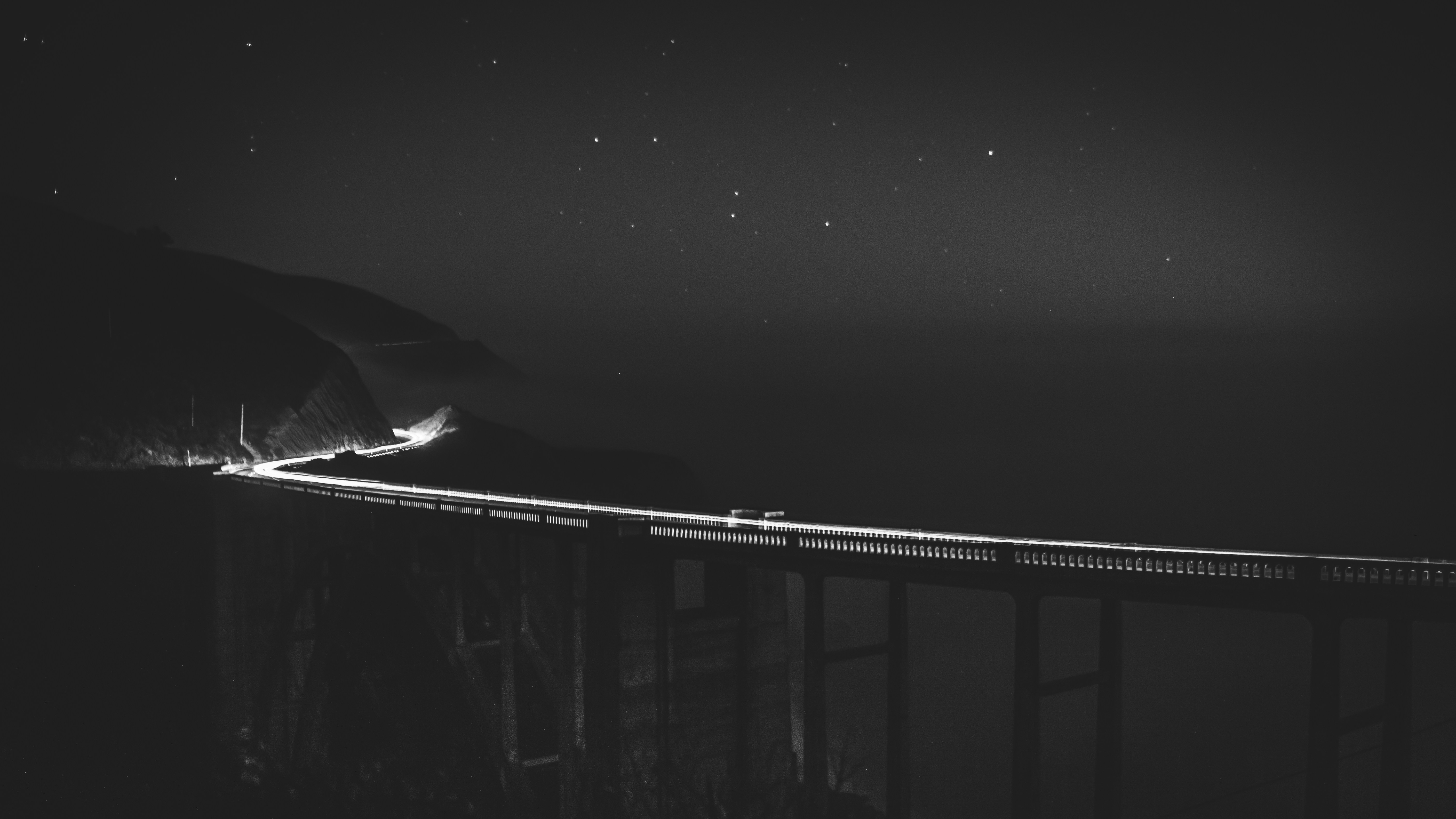 A winding bridge illuminated by vehicle headlights under a starry night sky, creating a serene contrast against the dark landscape.