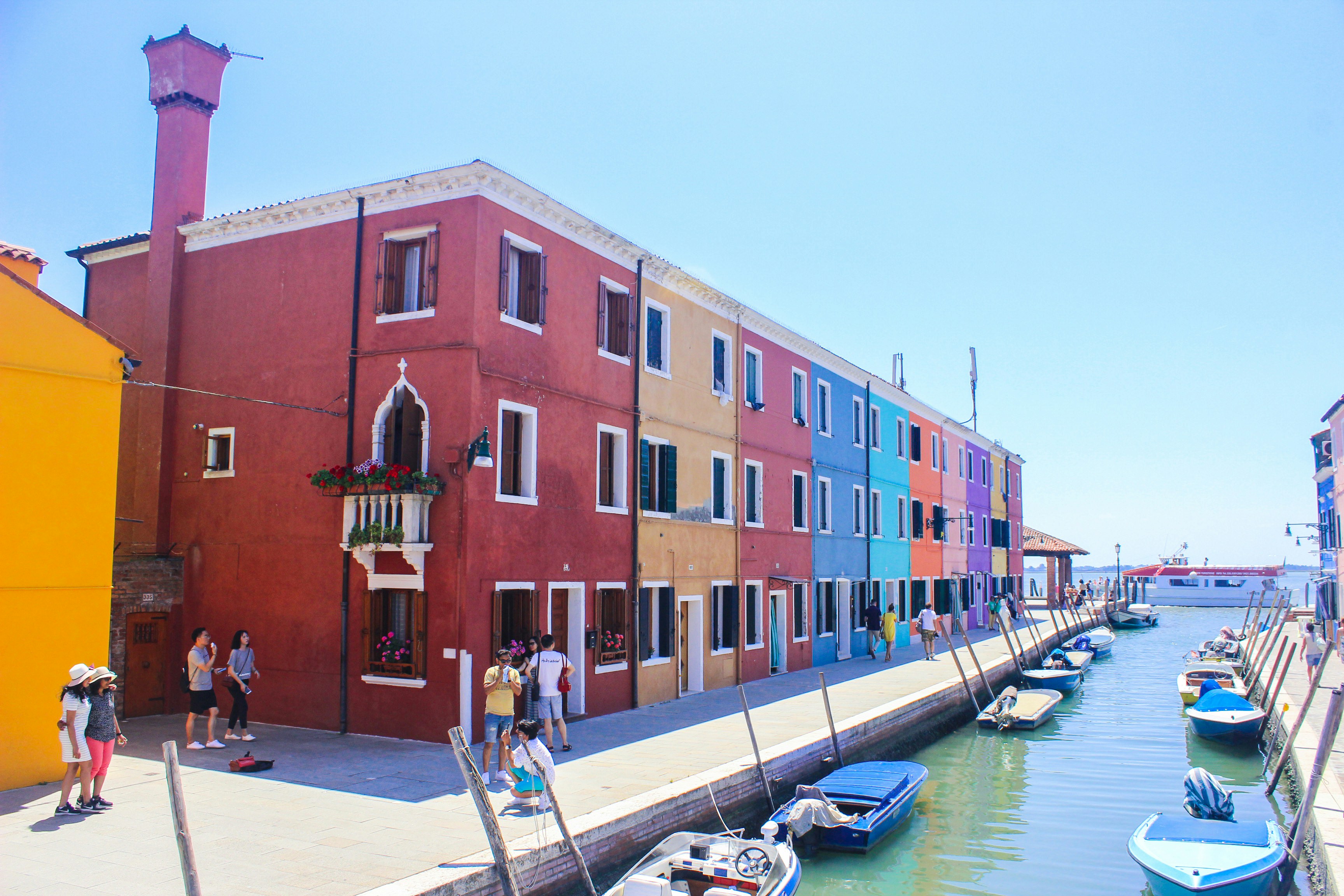 Colorful buildings line the canal in Burano, showcasing a lively atmosphere with people enjoying the sunny day. Boats are docked along the waterway.