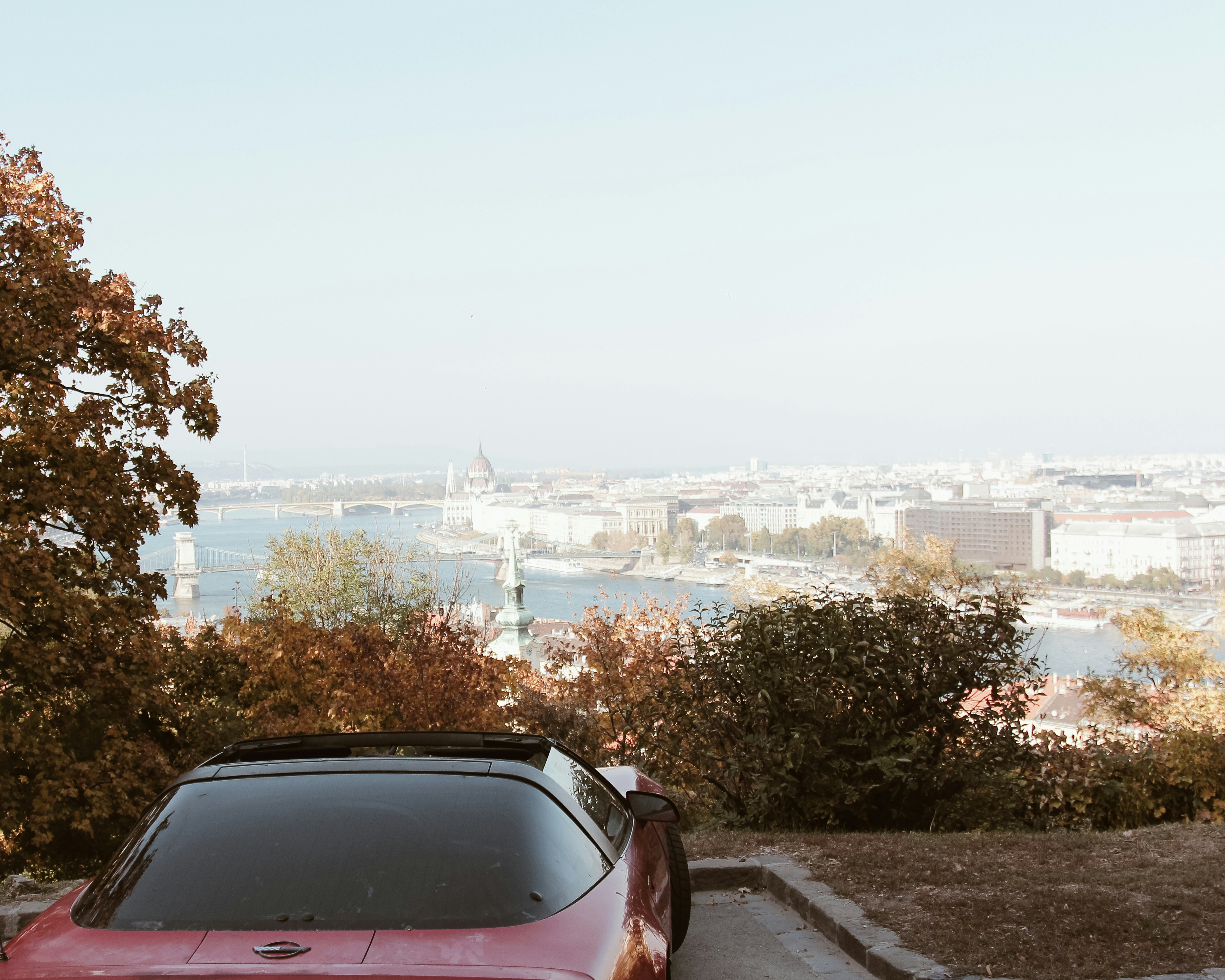 Red car parked on a hillside overlooking a vibrant cityscape with a river and distant landmarks. The scene is framed by autumn foliage.