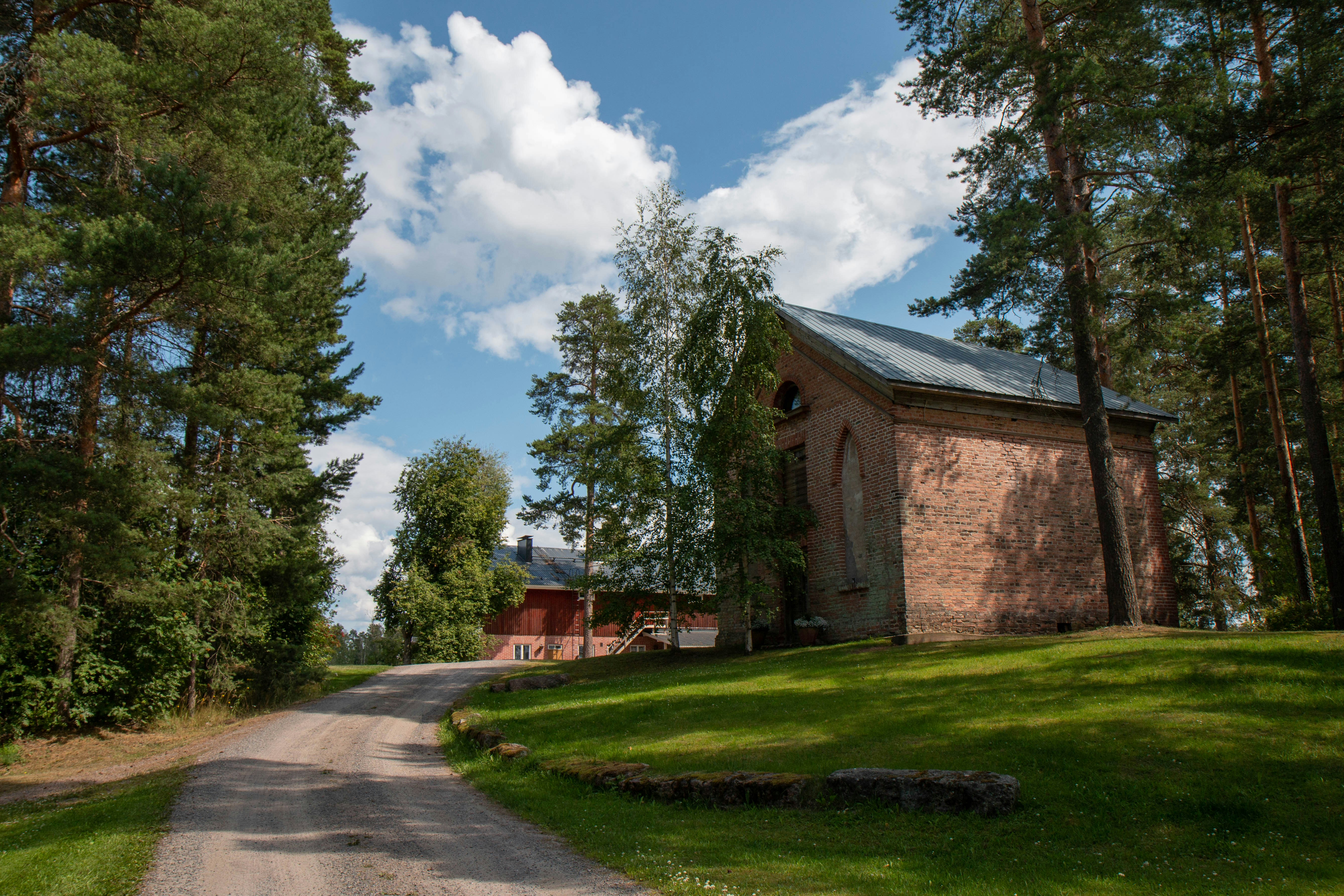 A rustic building nestled among tall trees, with a gravel path leading towards it under a bright blue sky dotted with clouds.