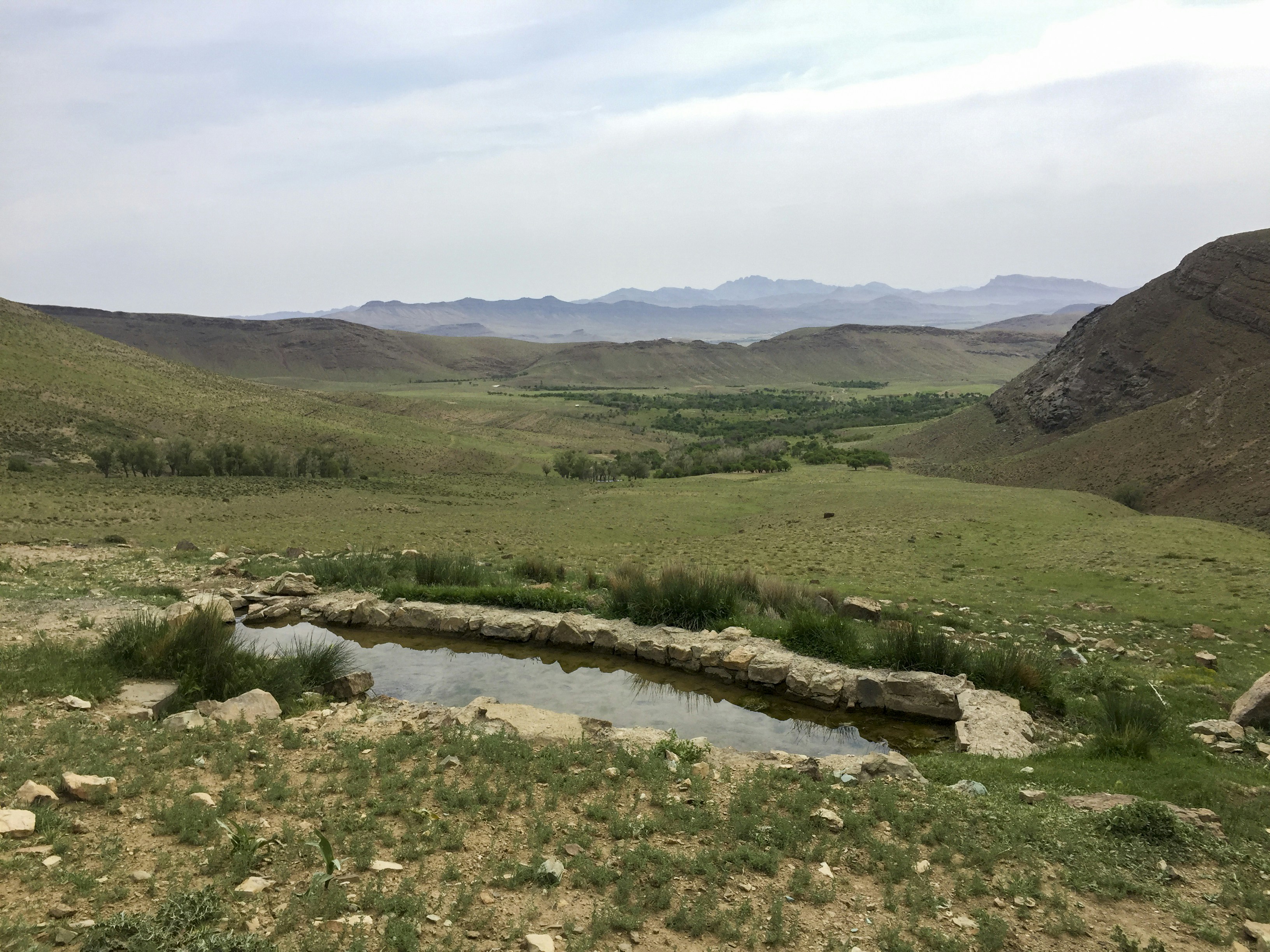 A tranquil stone pond surrounded by lush grasslands and distant mountains under a cloudy sky.