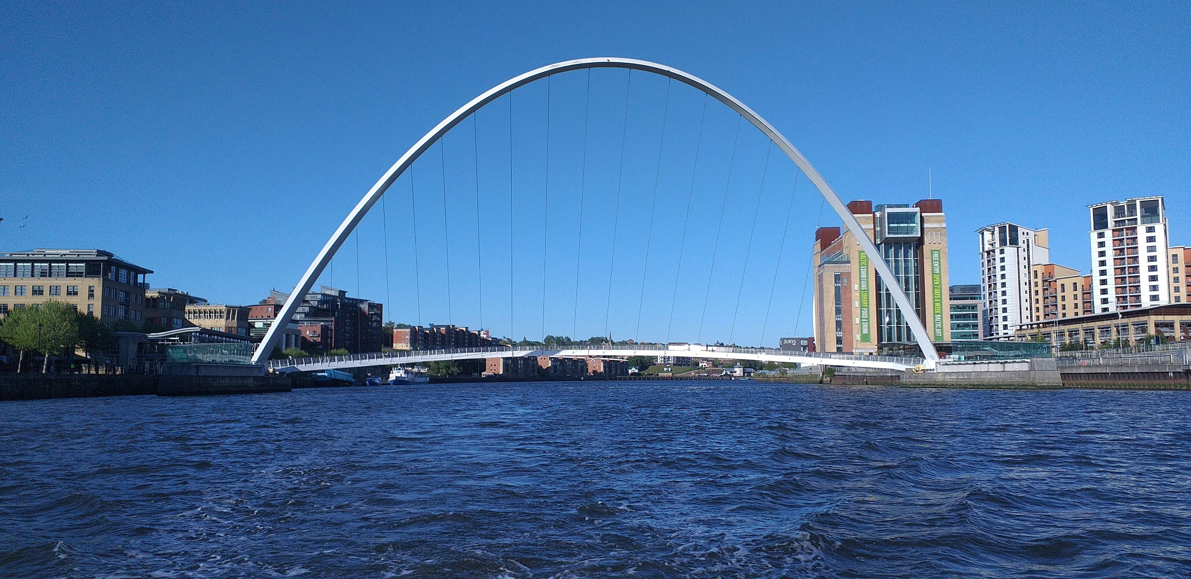 Elegant arched bridge spanning a wide river with modern buildings in the background under a clear blue sky.