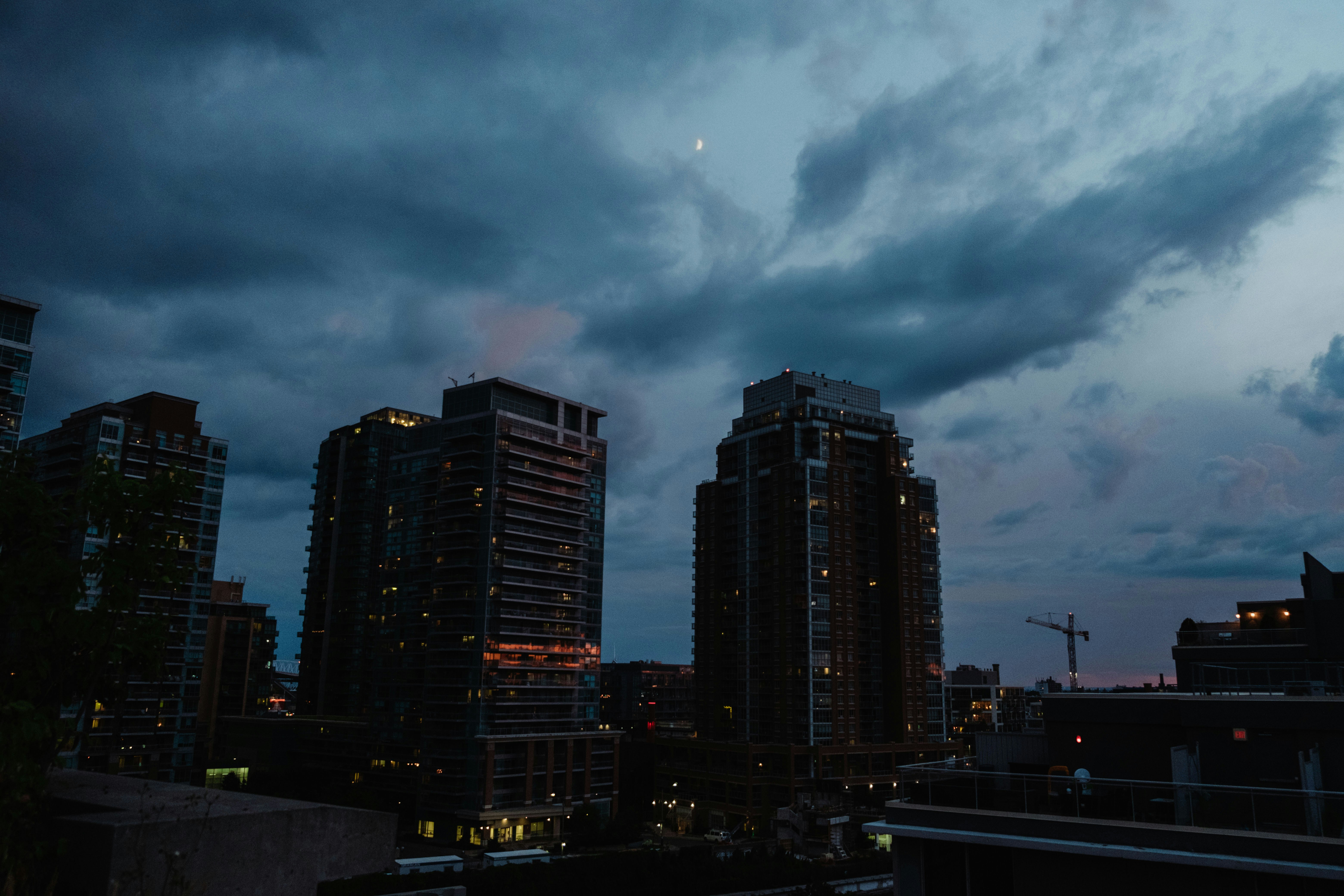 City skyline at dusk with towering buildings illuminated against a dramatic sky filled with clouds. A construction crane is visible in the background.
