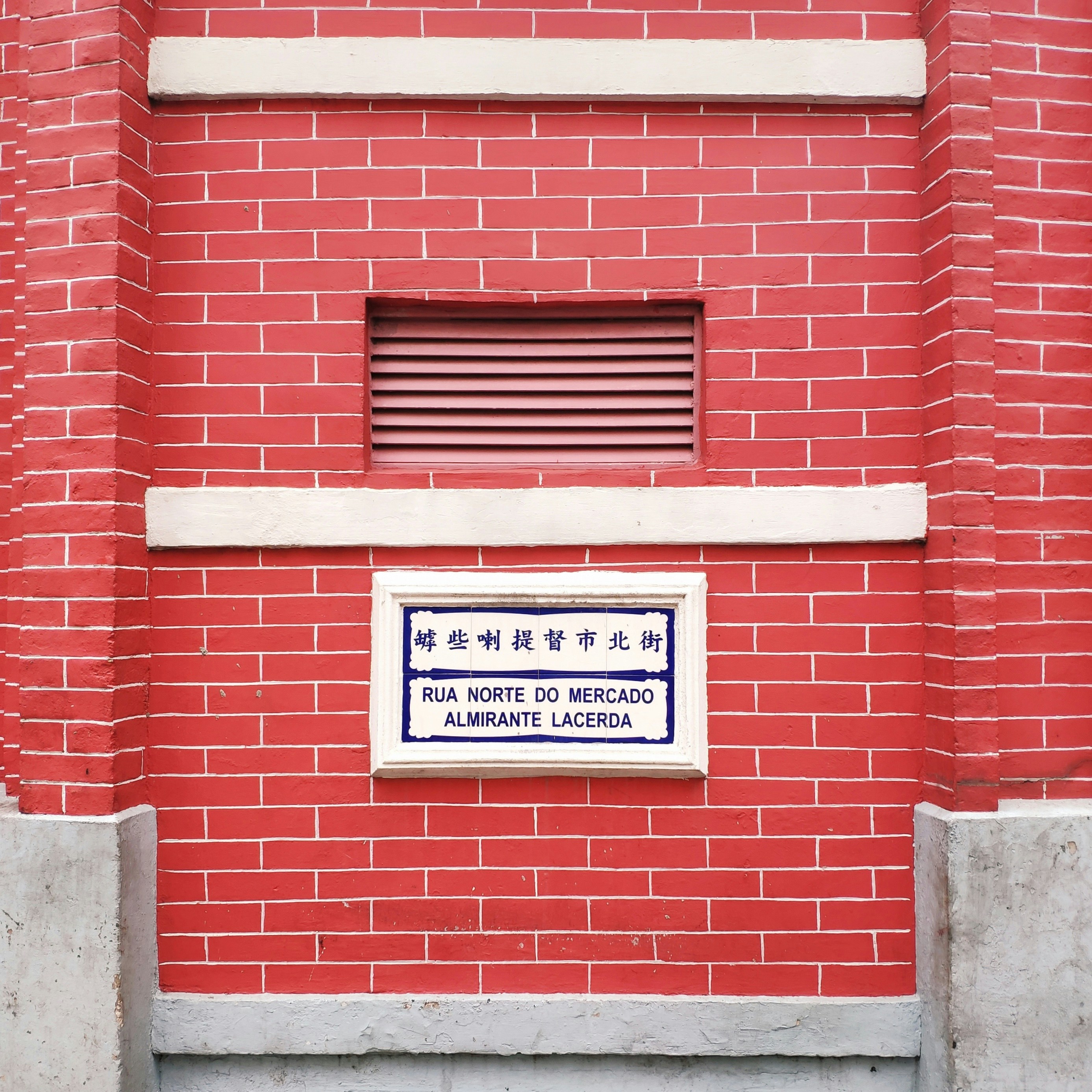 Photo of a red brick wall featuring a vent above a blue-and-white street sign framed by concrete ledges.