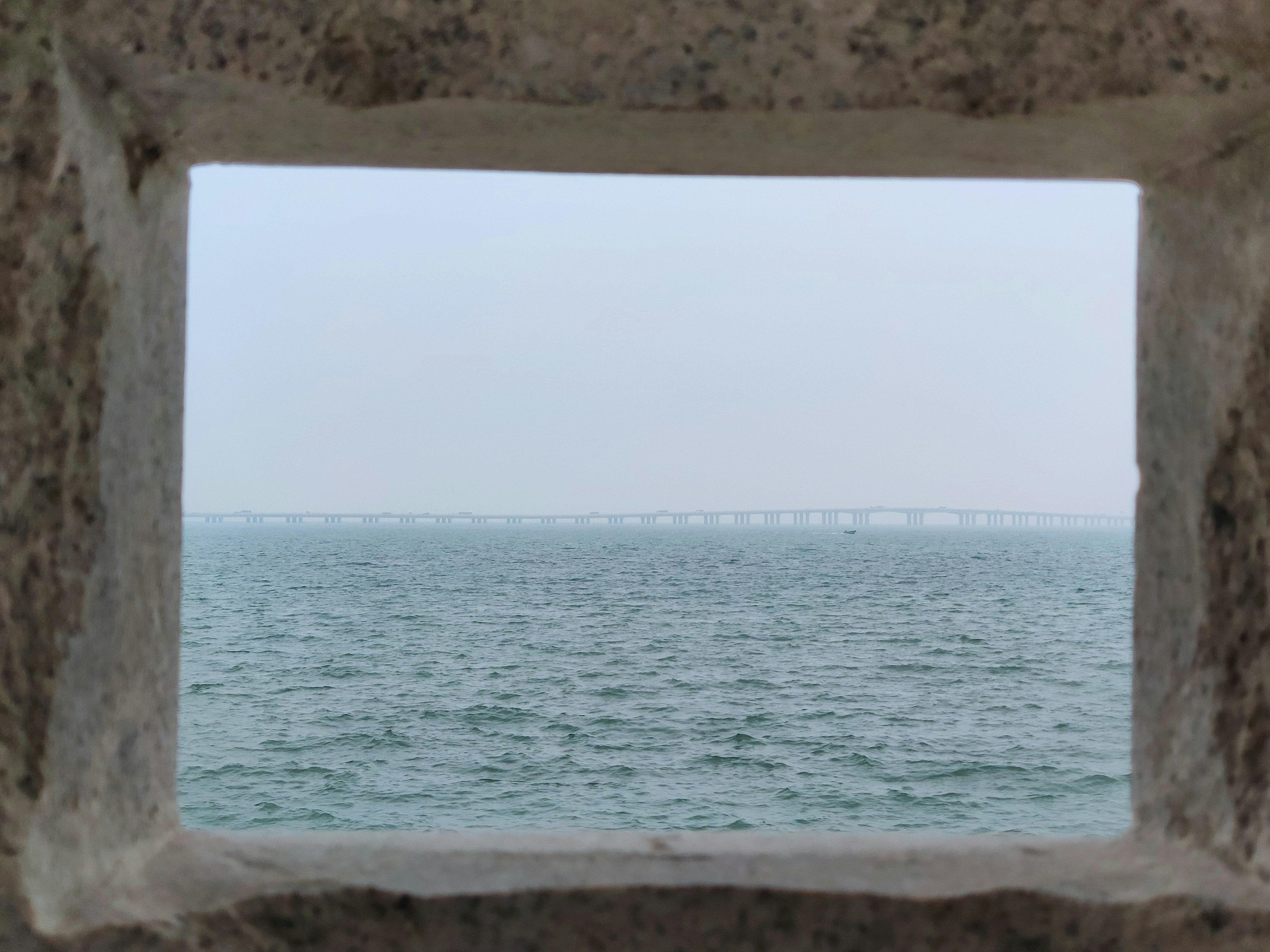 View of the ocean framed by a stone window with a distant bridge on the horizon.