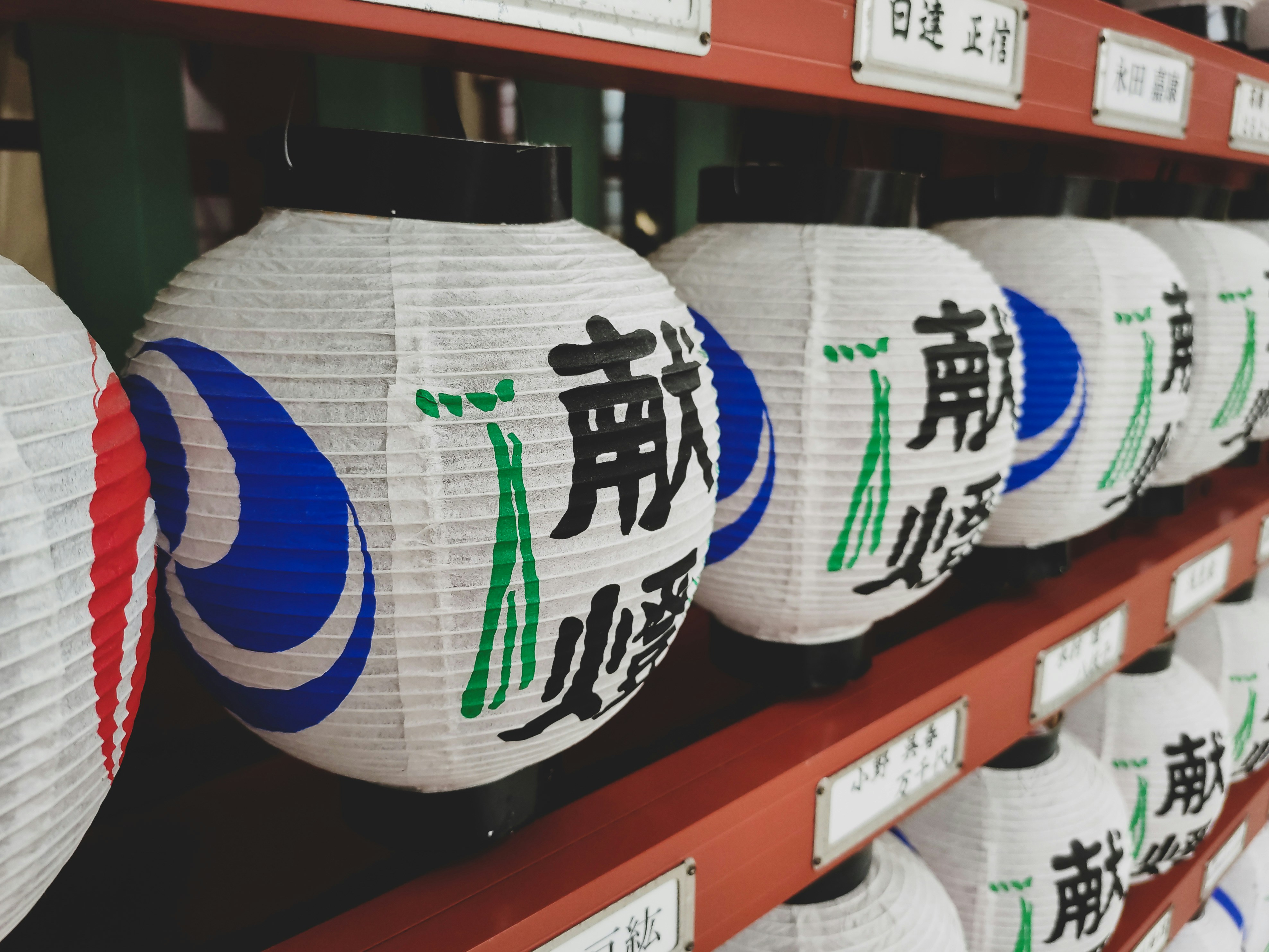 Close-up photograph of white paper lanterns with bold black kanji and green accents, arranged on a red market shelf.