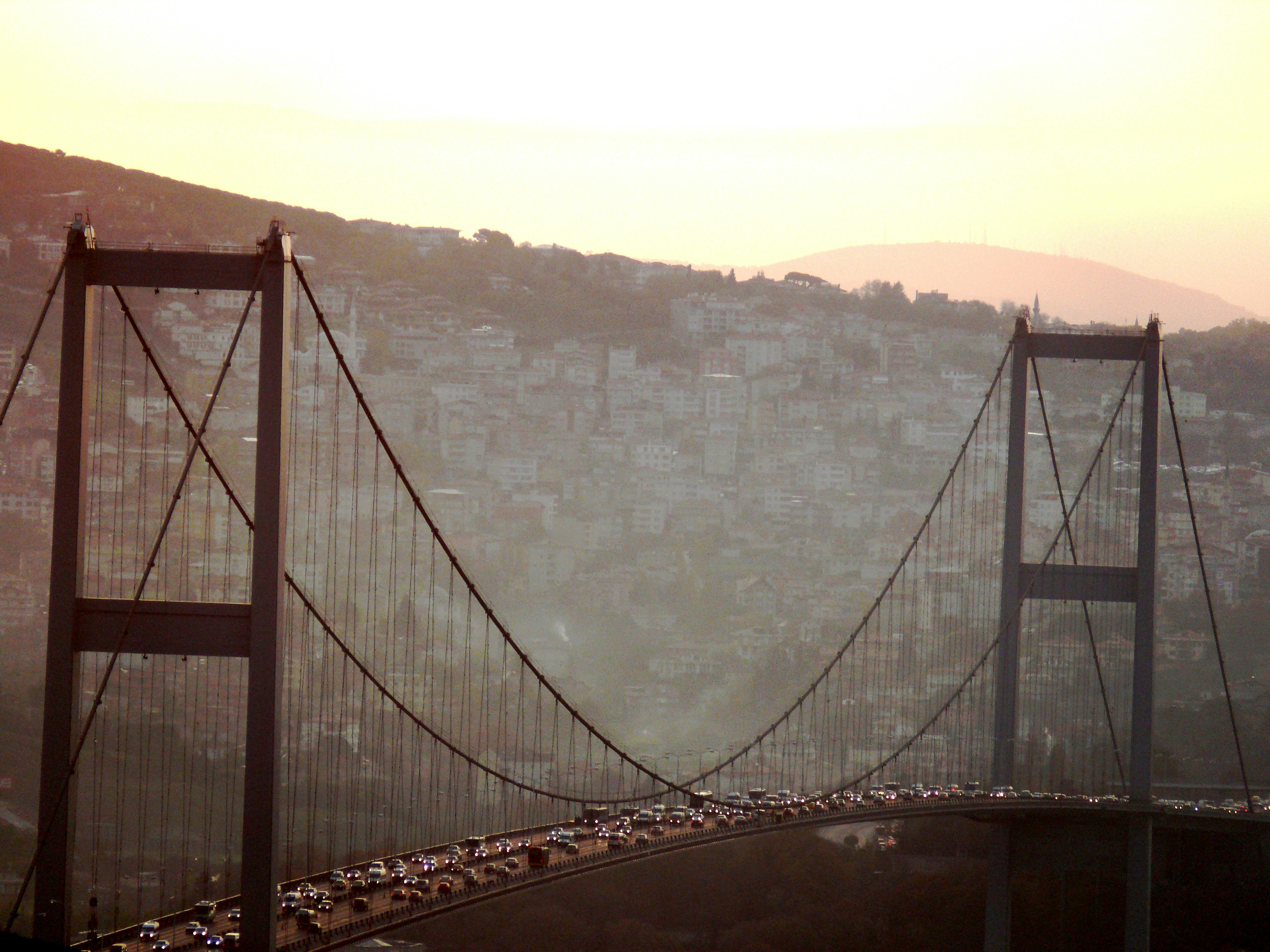 Traffic flows over the Bosphorus Bridge with a hazy cityscape backdrop at sunset.