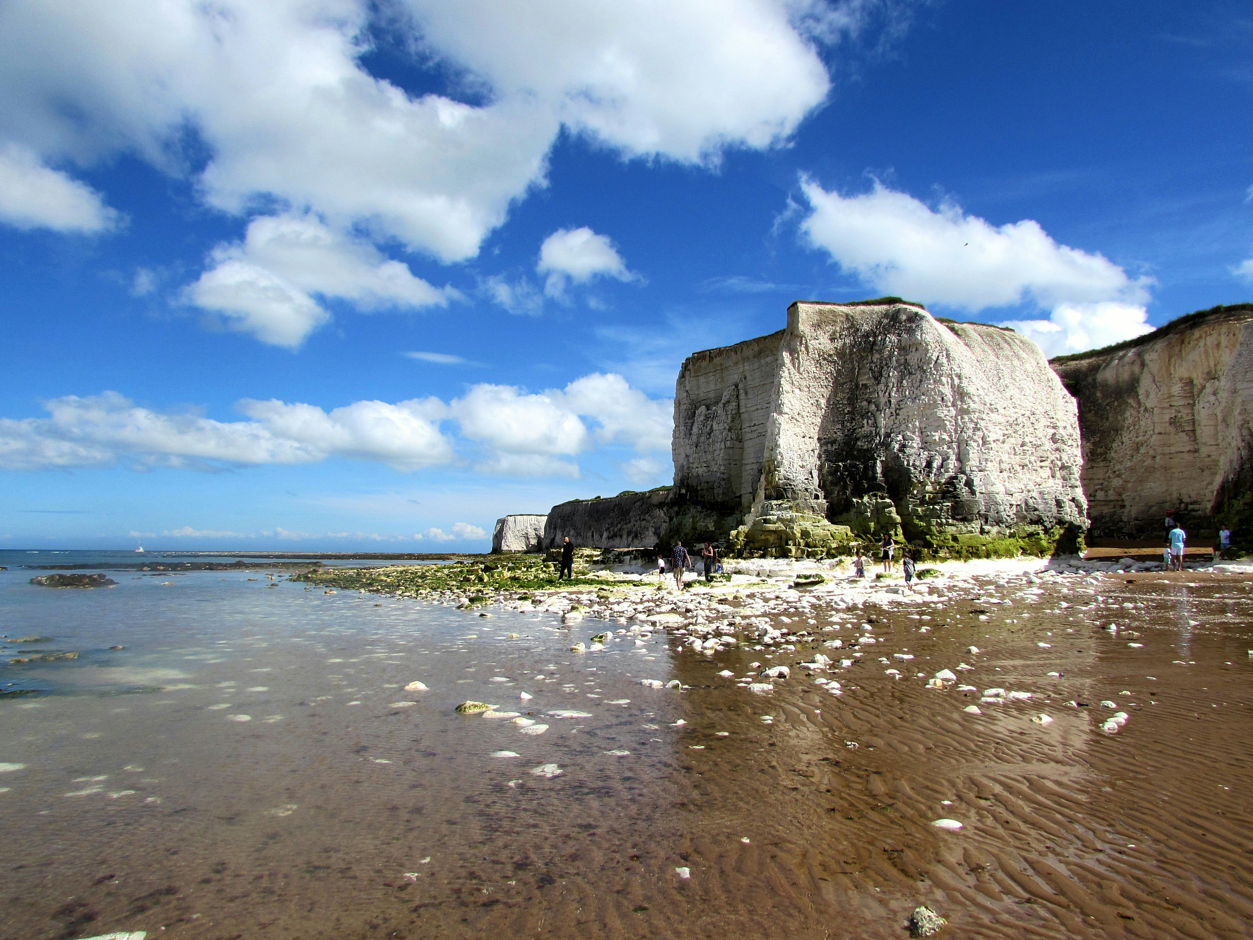 Weathered cliffs rise beside a shallow tide pool under scattered clouds and a vibrant blue sky.