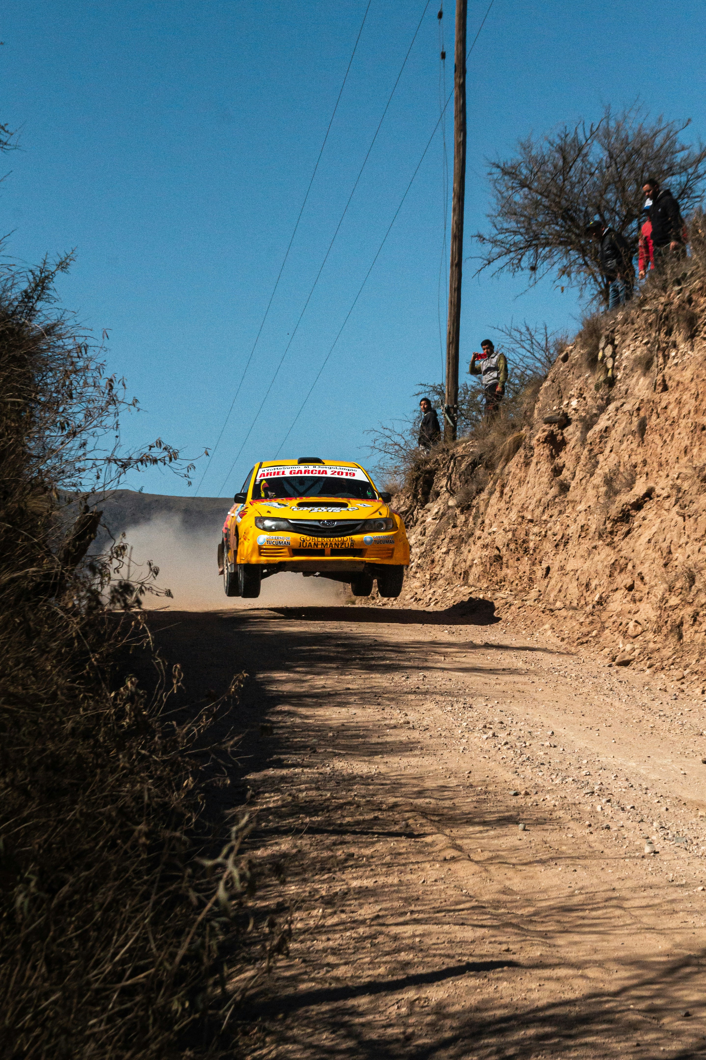 Yellow rally car airborne on a dusty road under clear blue sky.