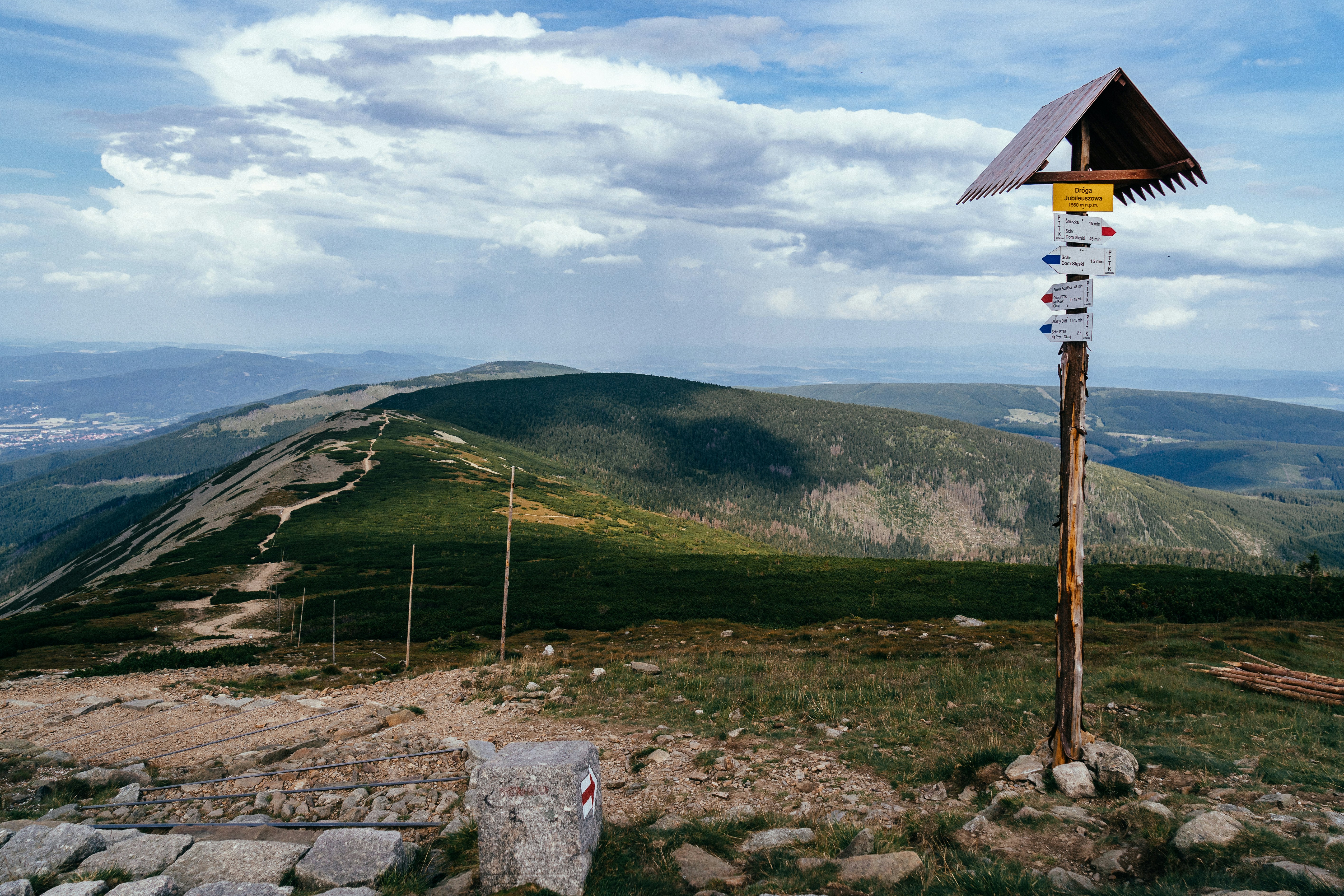 A wooden signpost marks the beginning of a hiking trail on a verdant mountain slope, with expansive views of the rolling hills and distant valleys under a partly cloudy sky.
