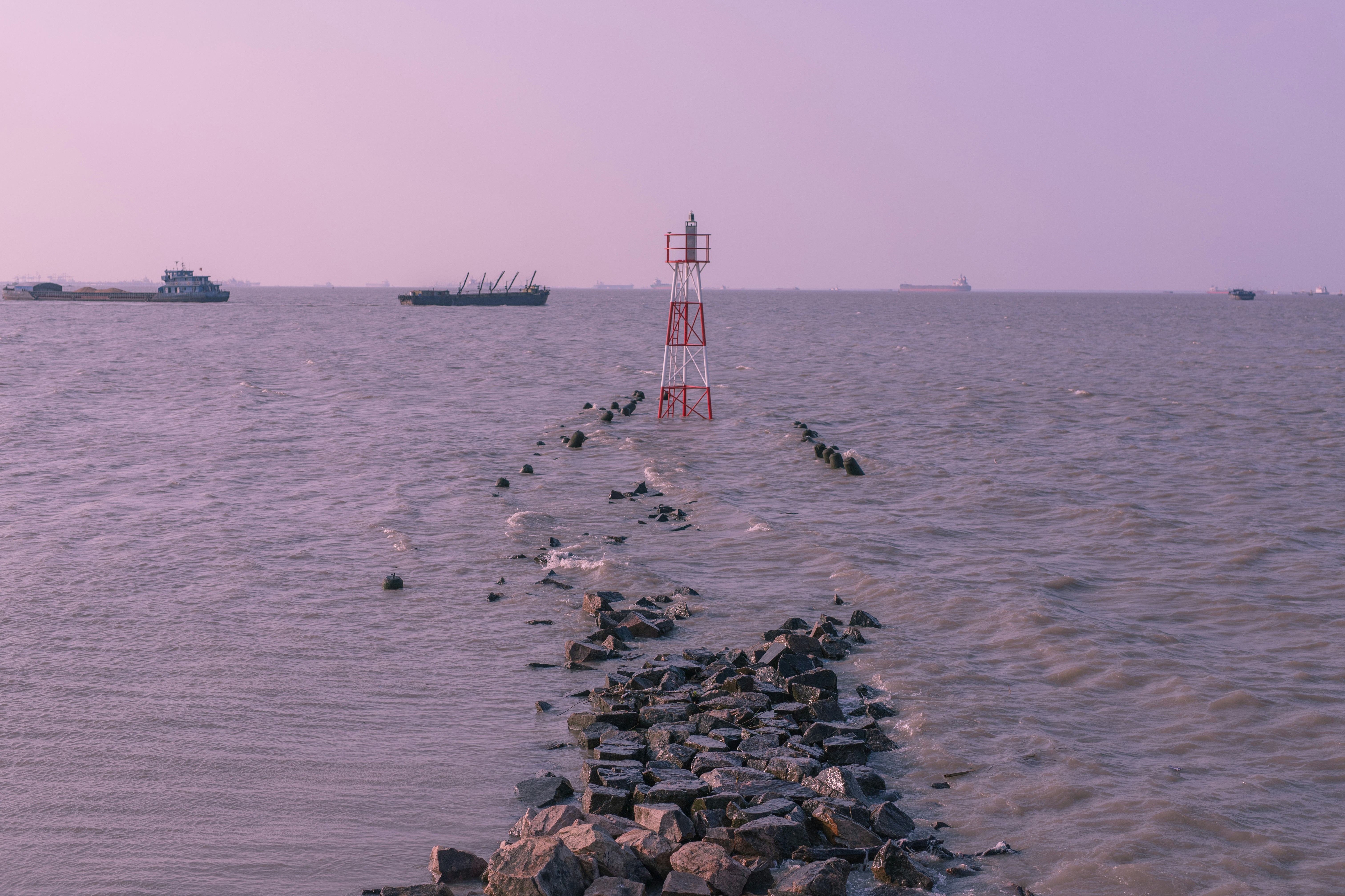 A red and white lighthouse stands tall at the edge of a rocky jetty, guiding vessels through calm waters under a pastel sky.